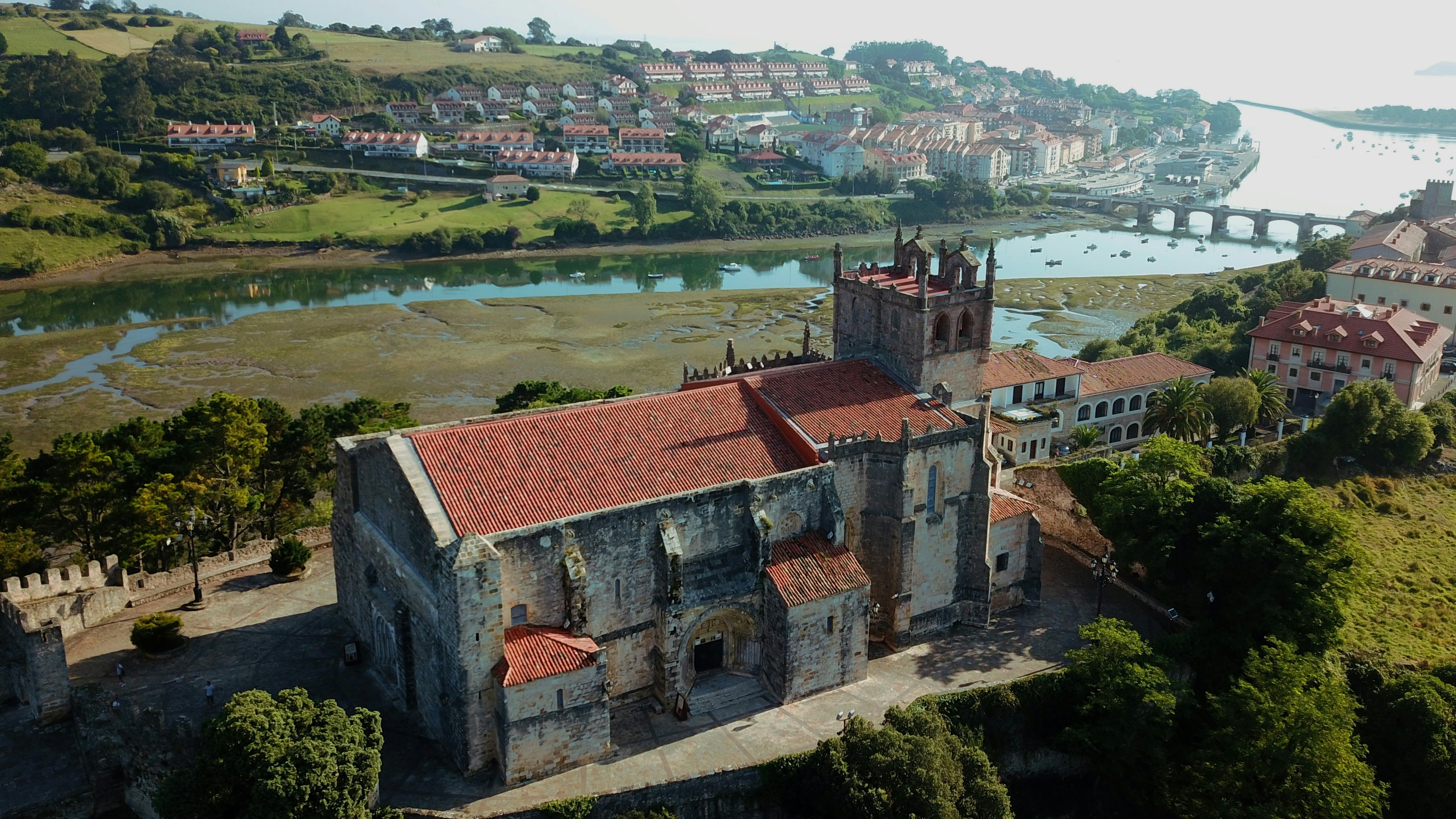 Historic monastery with a red-tiled roof situated near a serene river, surrounded by lush greenery and modern housing in the background.