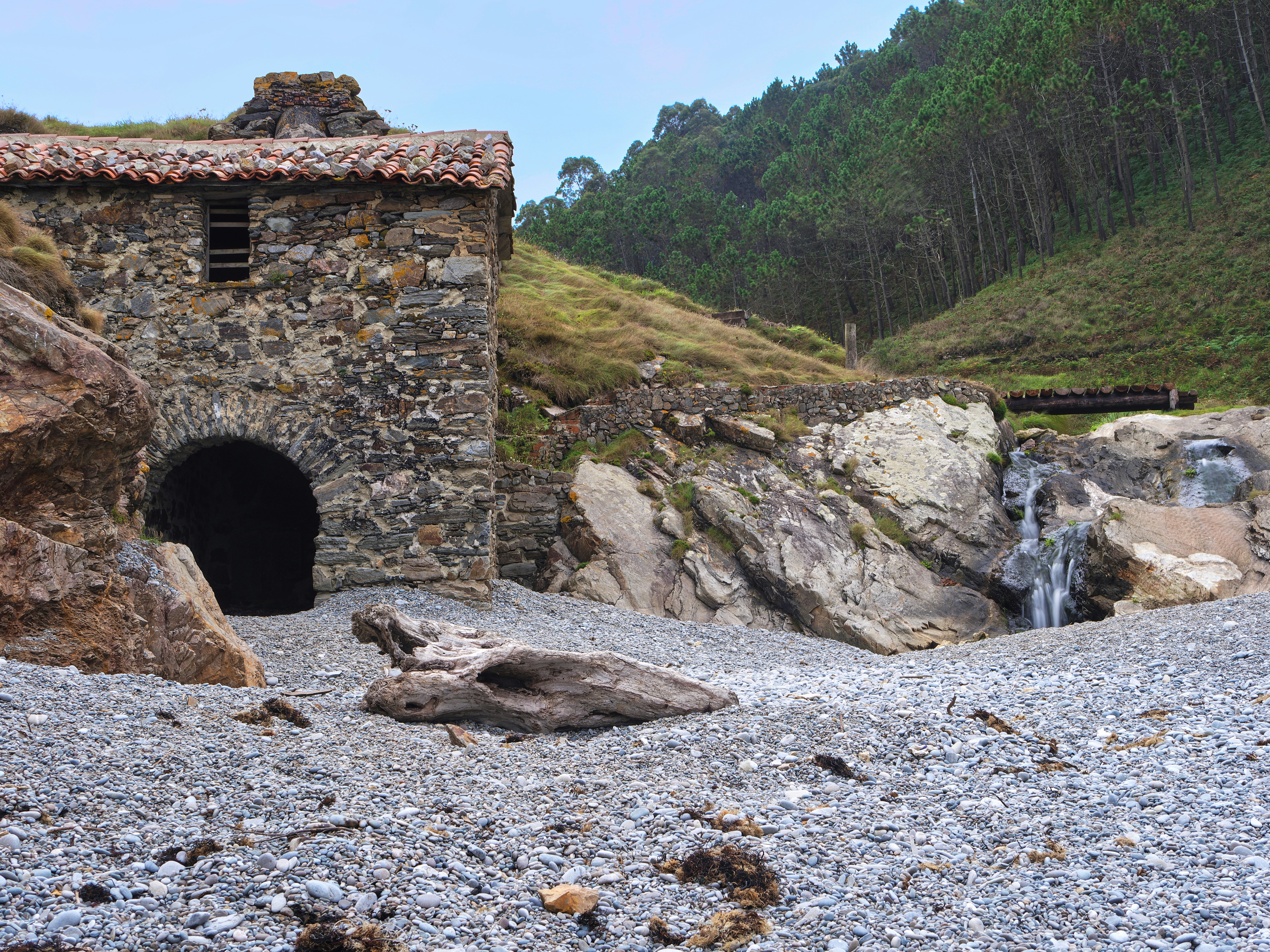 Weathered stone structure nestled among rocky terrain and lush greenery, hinting at a forgotten history. A gentle waterfall cascades nearby.