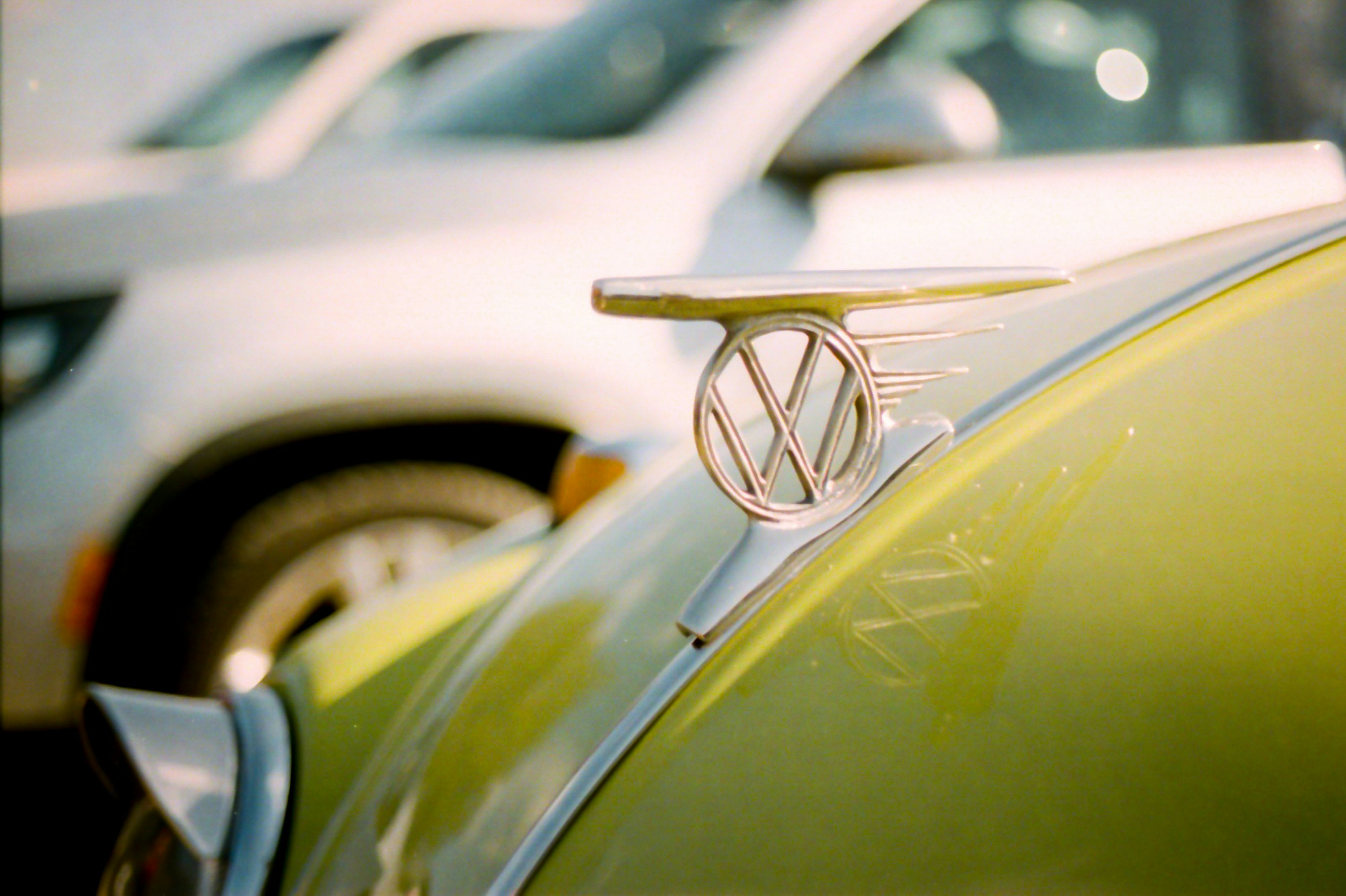 Close-up of a vintage chrome hood ornament logo on a lime-green car, with a softly blurred background.