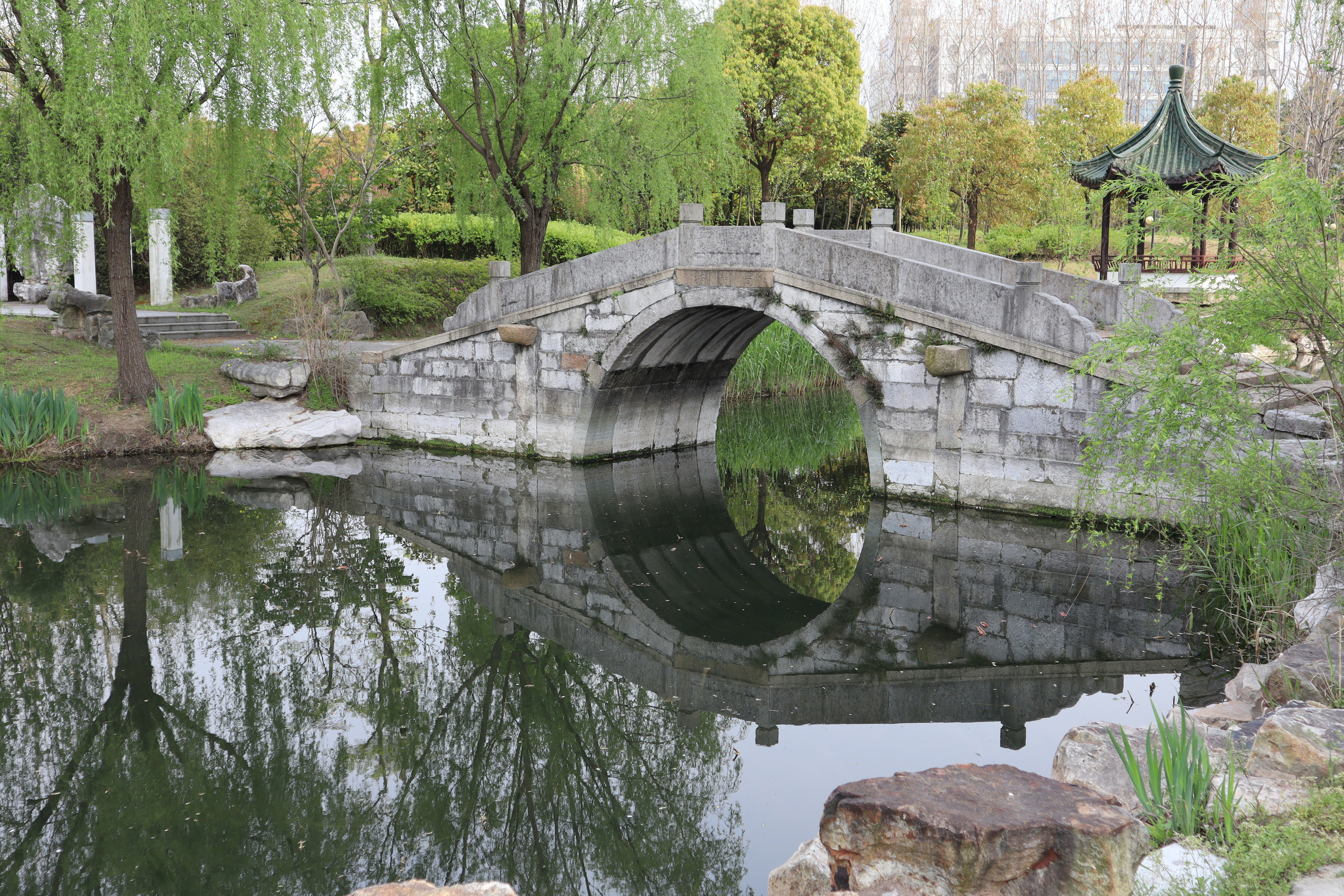 Stone arch bridge reflected in calm waters of a serene park, surrounded by lush greenery and a traditional gazebo.