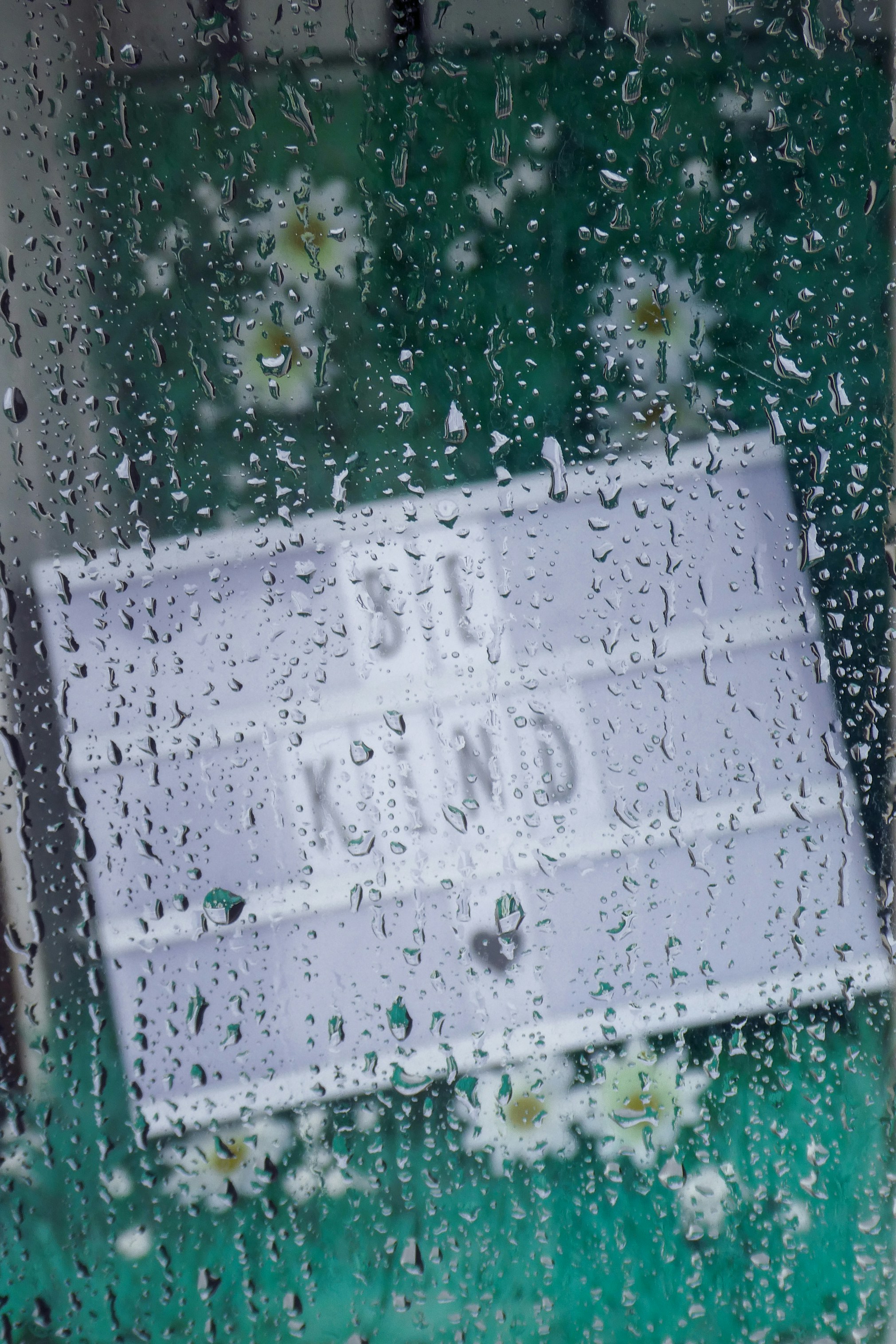 Rain droplets cling to a glass pane, obscuring a white sign behind a green frame. This photograph highlights the texture of water beads on glass and the blurred urban signage beyond.