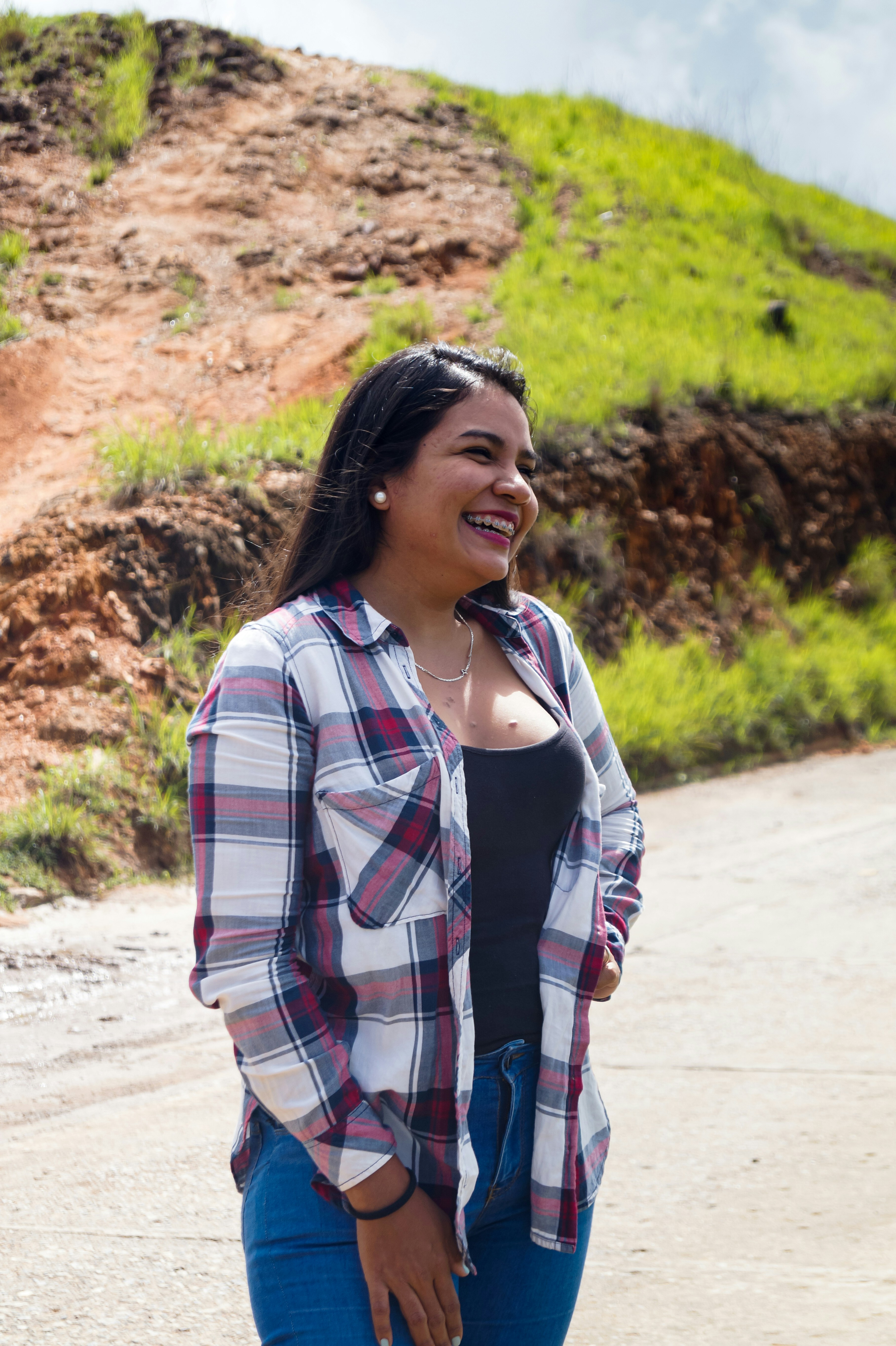 Smiling woman in a checkered shirt stands on a sunlit path with grassy hills in the background.