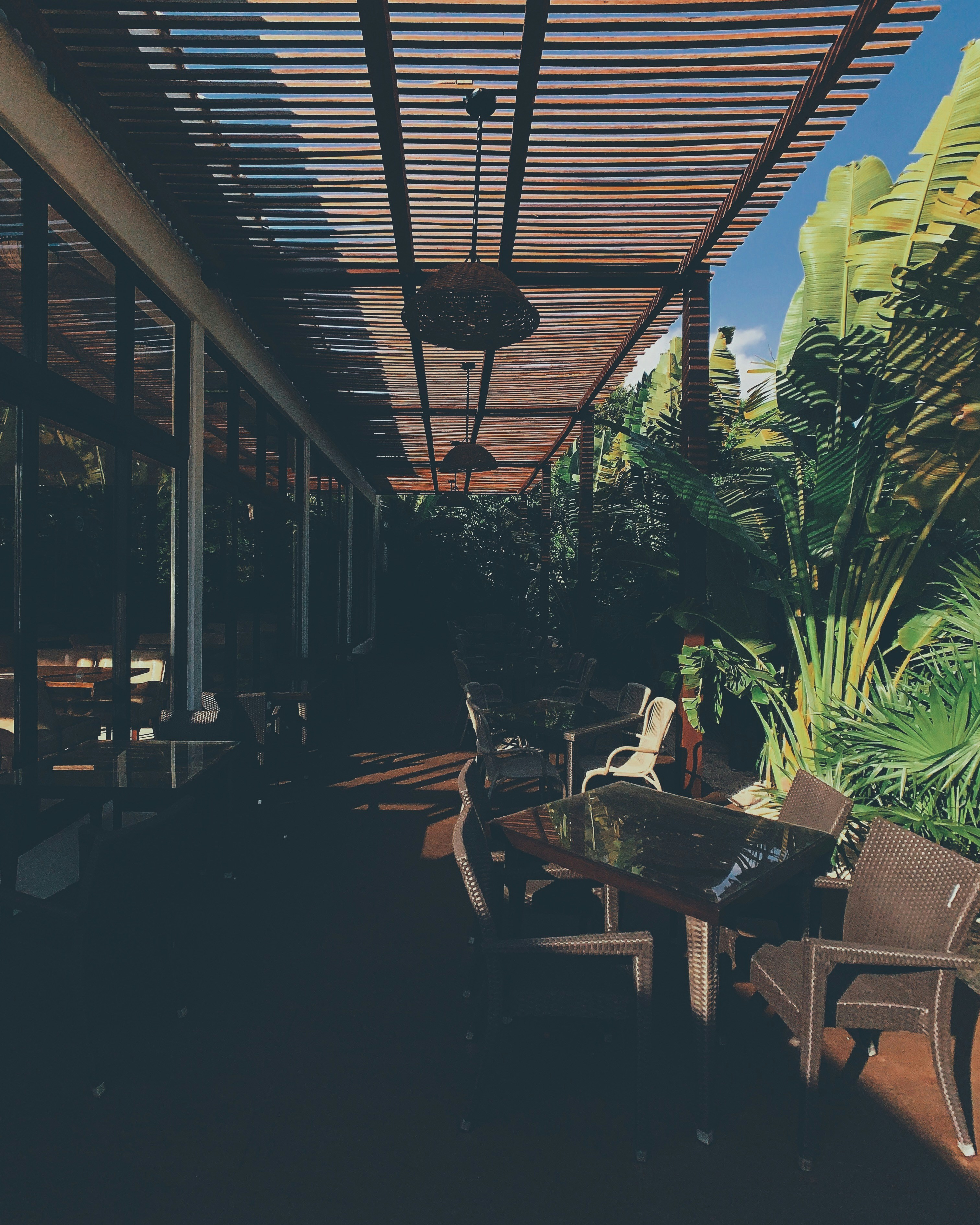 A cozy outdoor dining area shaded by wooden slats, surrounded by lush greenery and a bright blue sky.