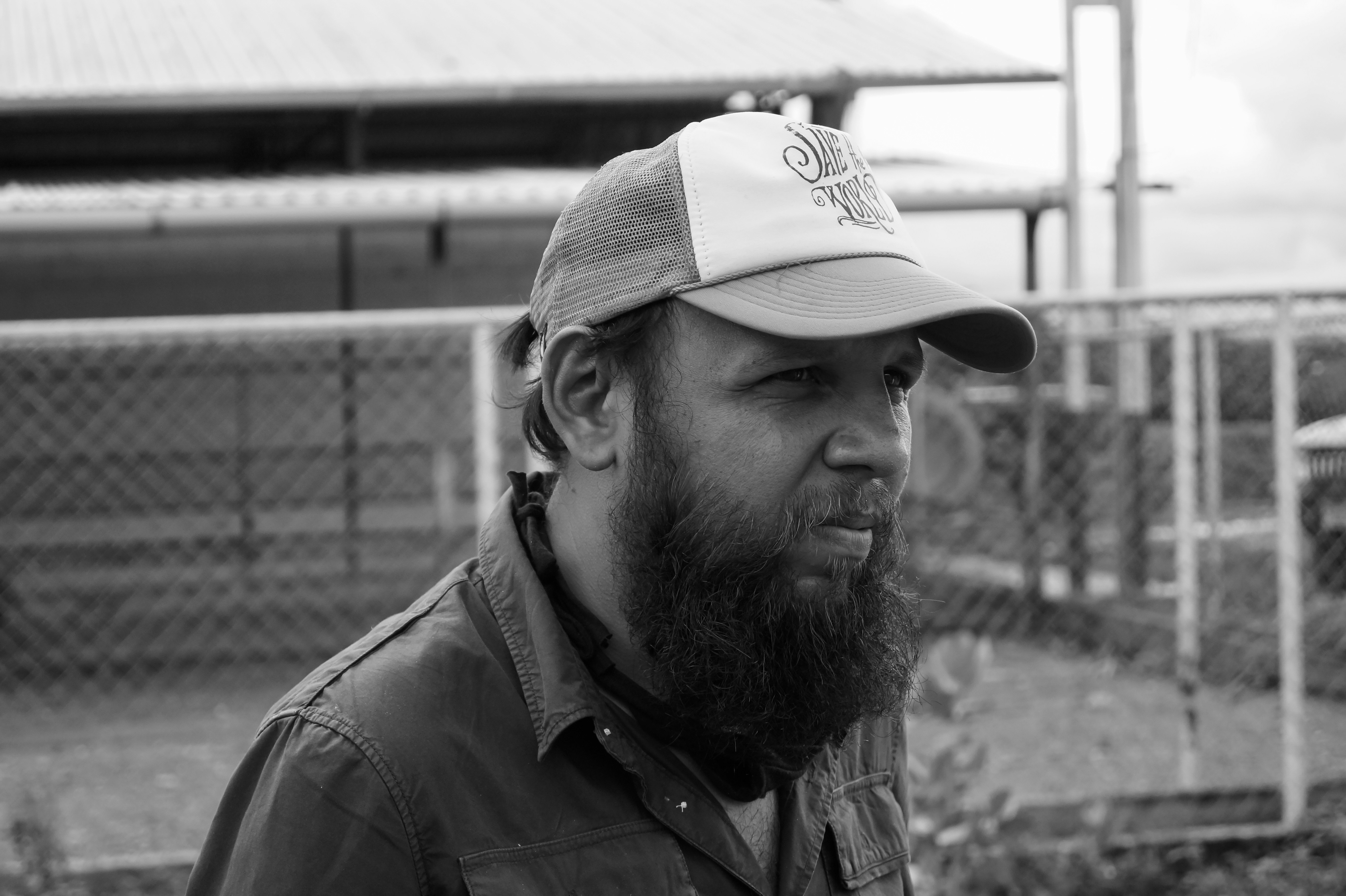 Black and white portrait of a bearded man in a cap, standing near a fence with a contemplative expression.