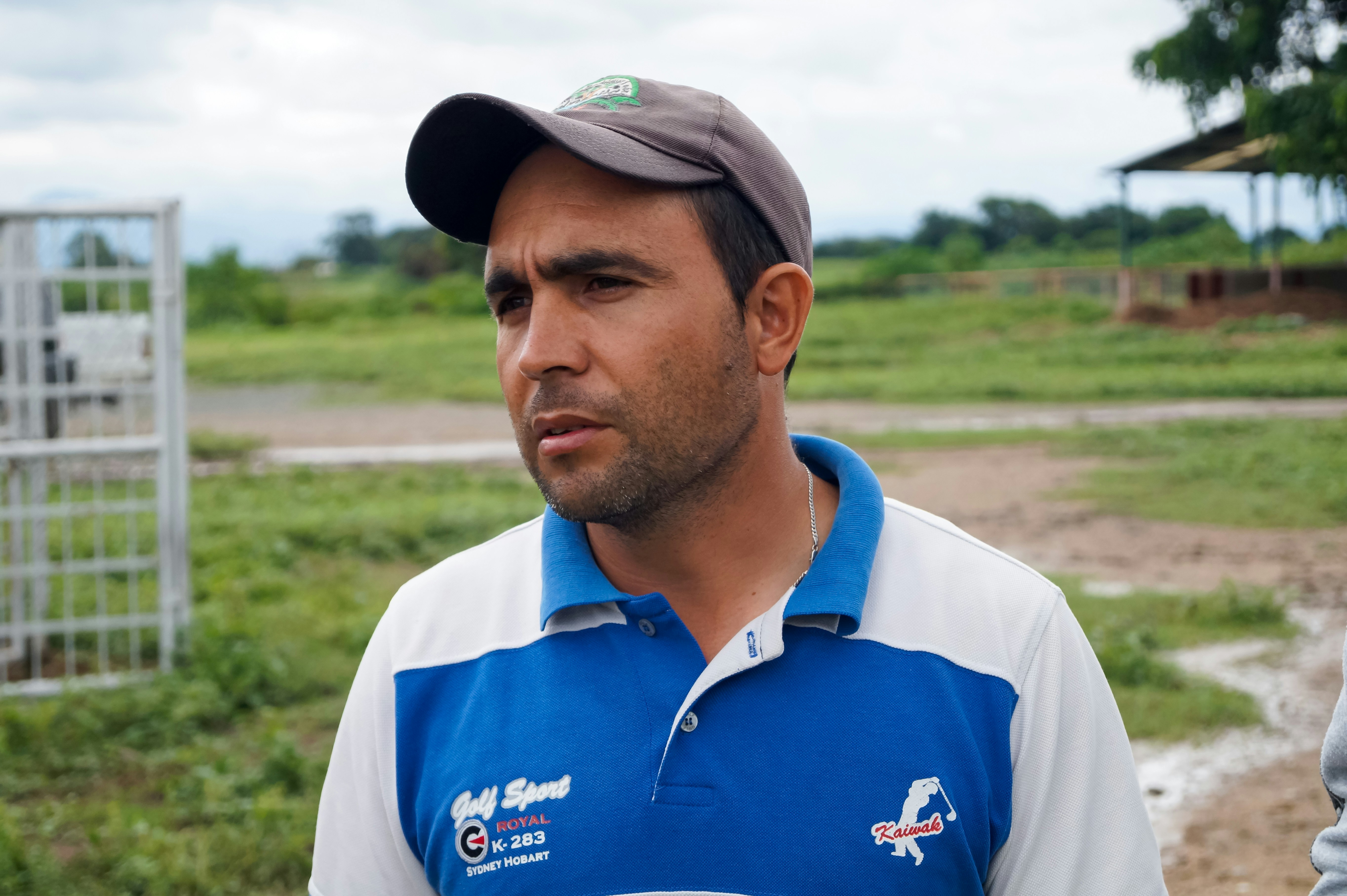 Man in a blue and white shirt stands outdoors with a thoughtful expression, surrounded by greenery and cloudy skies.