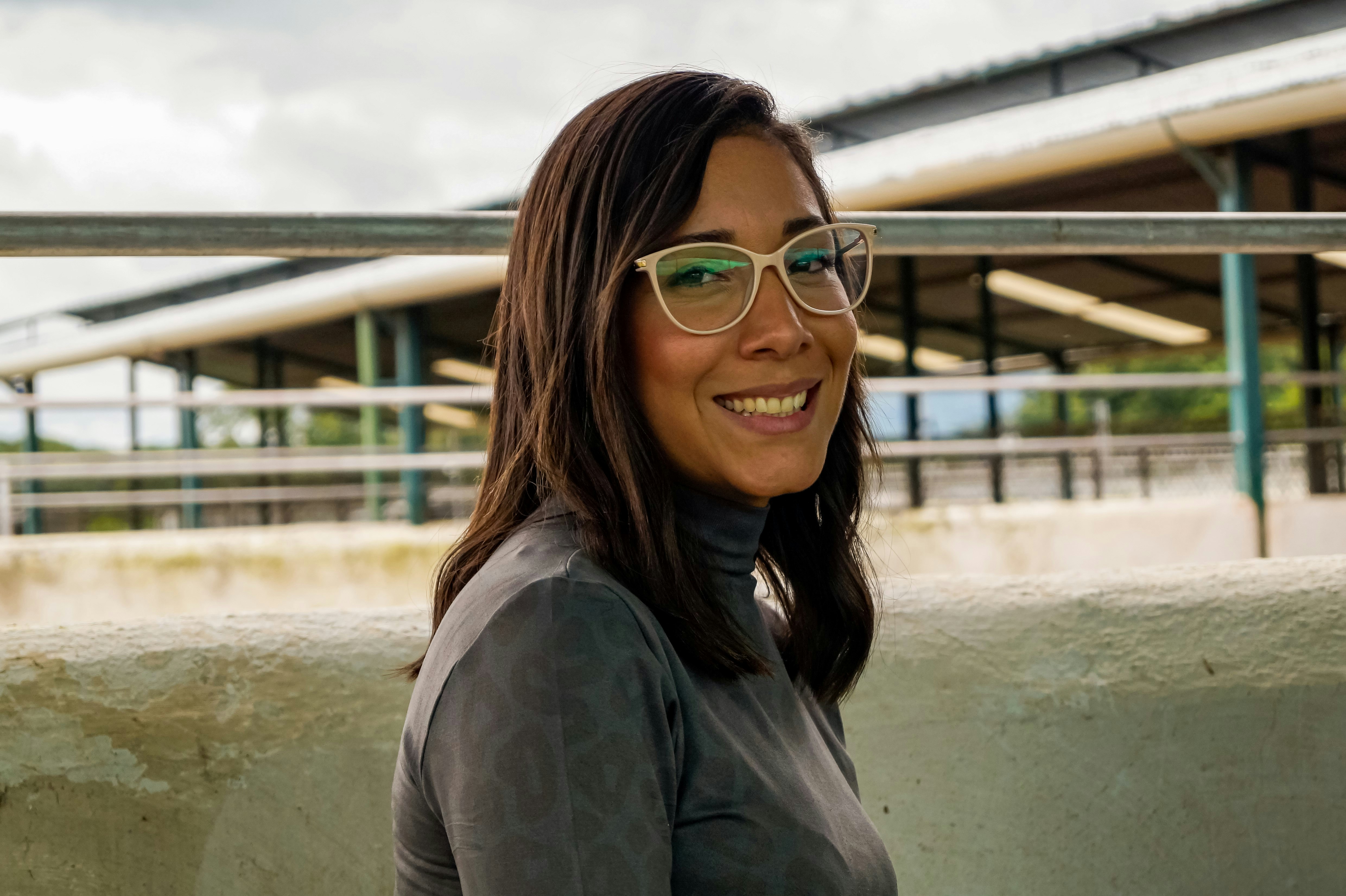 Smiling person wearing glasses with an industrial backdrop under a cloudy sky.