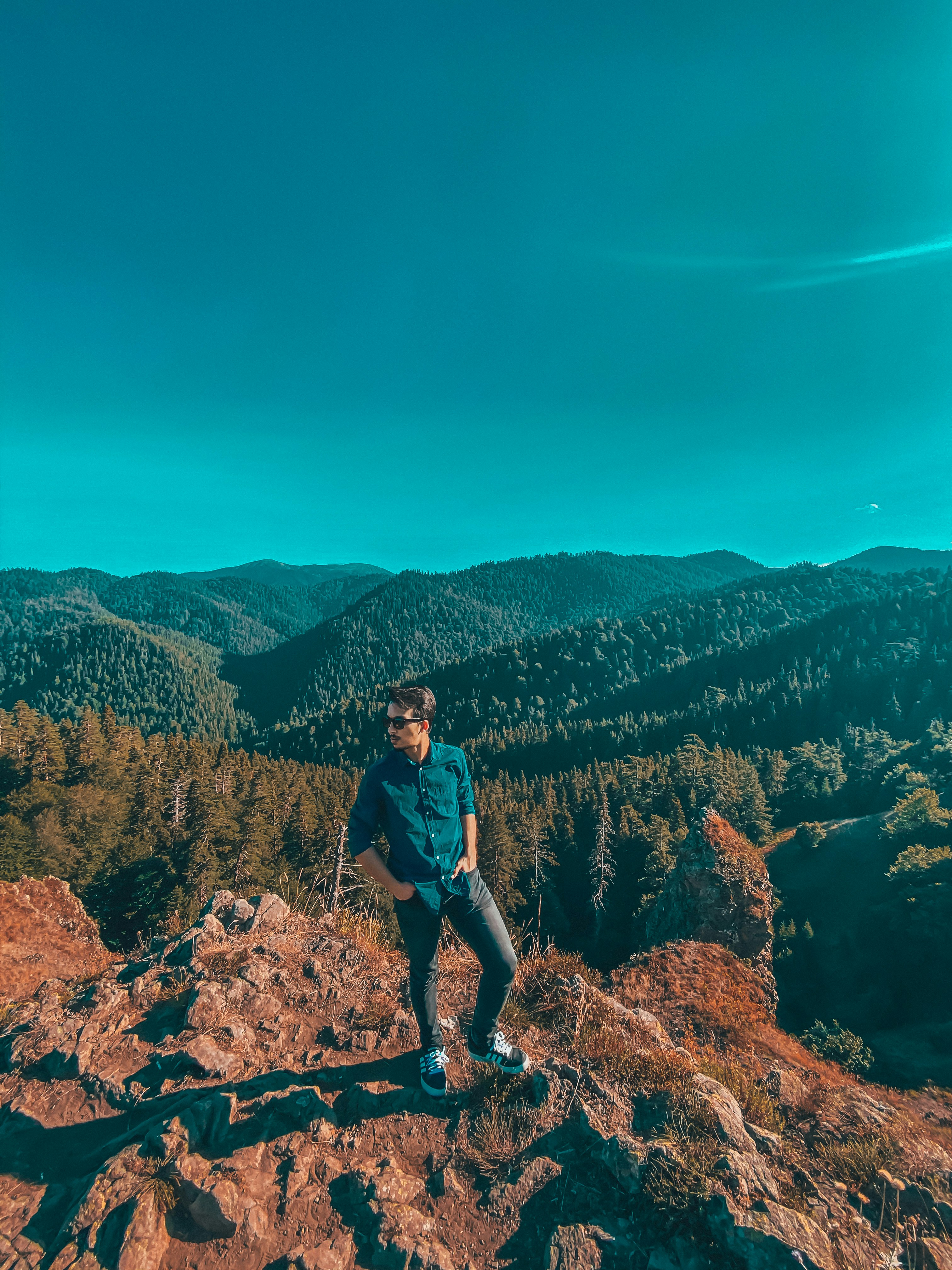 A person stands confidently on a rocky outcrop, overlooking a vast expanse of forested mountains under a clear blue sky.