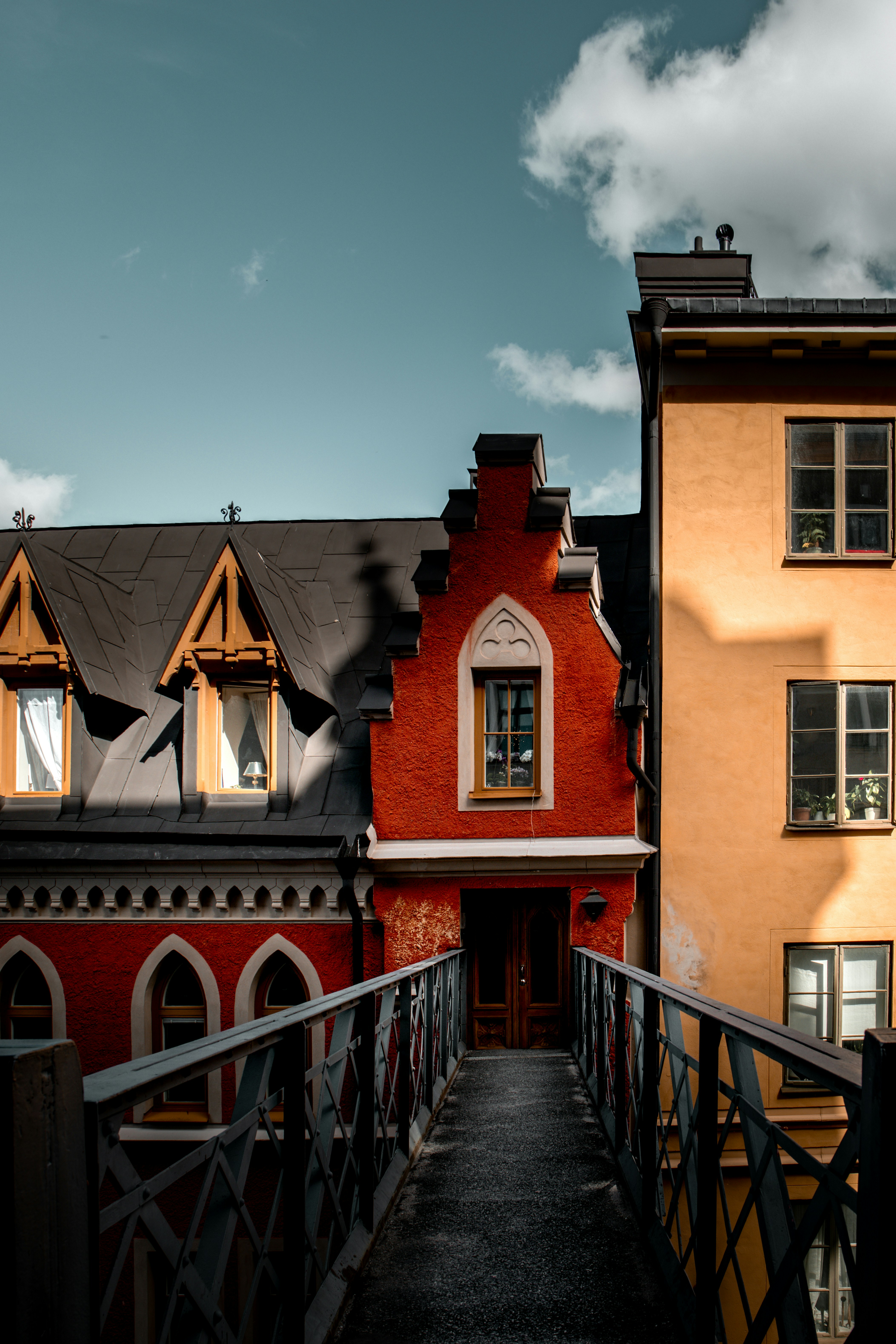A vibrant red building with intricate architectural details stands alongside a yellow structure, connected by a narrow bridge. The interplay of light and shadow enhances the scene's character.