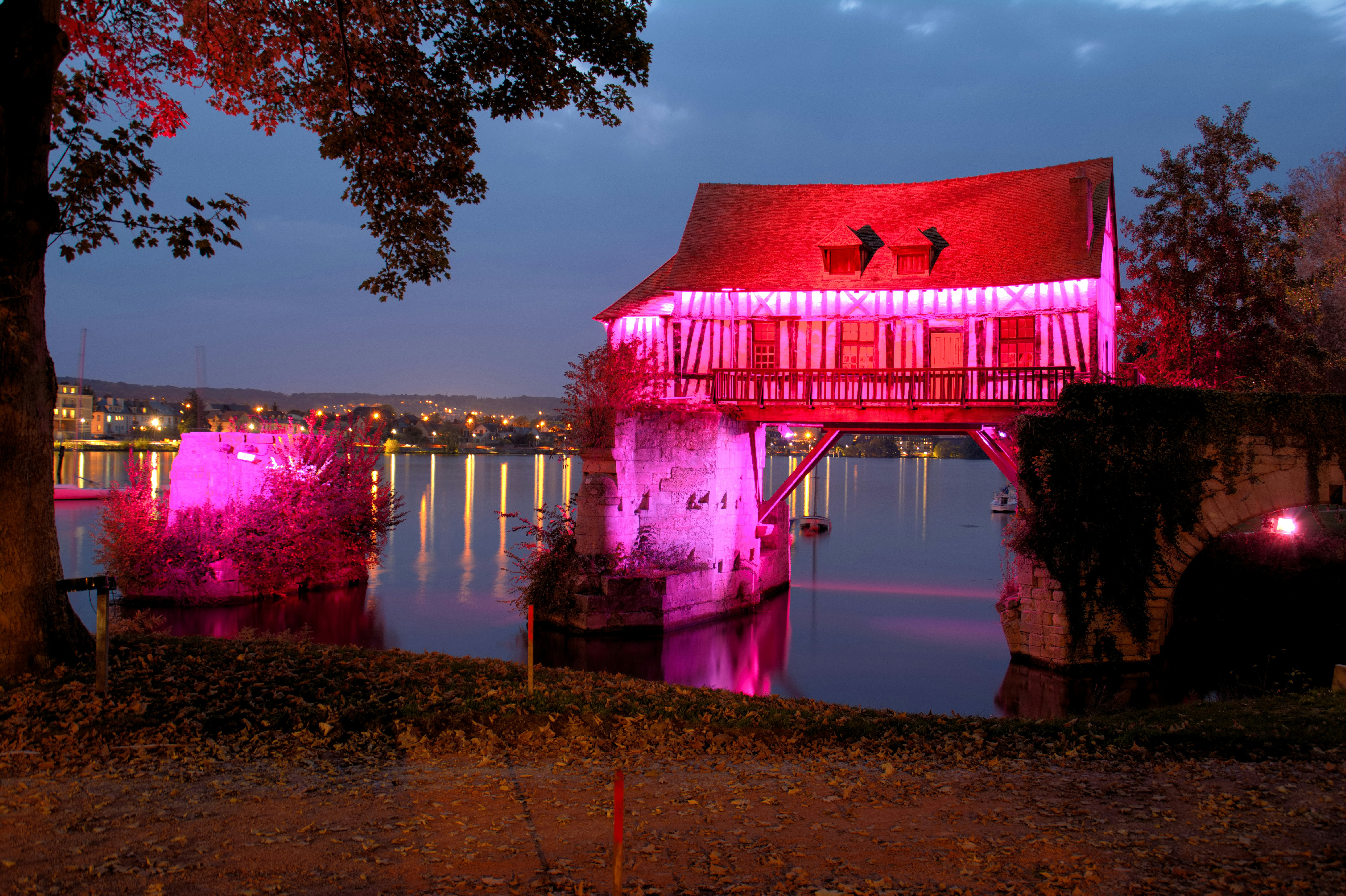 Historic mill building glowing with pink lights over a tranquil river at dusk.