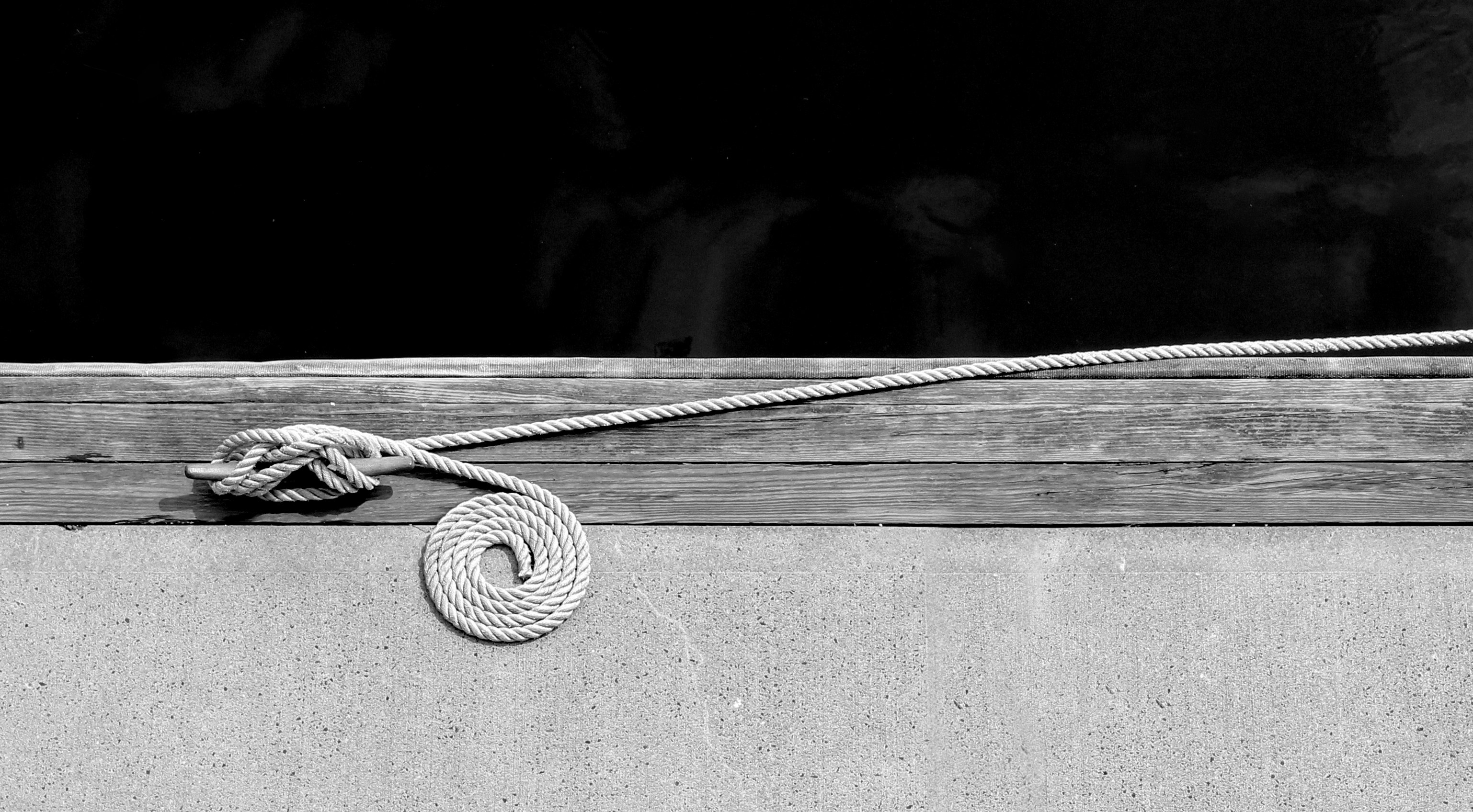 A coiled rope resting against a wooden dock, contrasting with the dark water below. The composition highlights the textures and shapes of maritime elements.