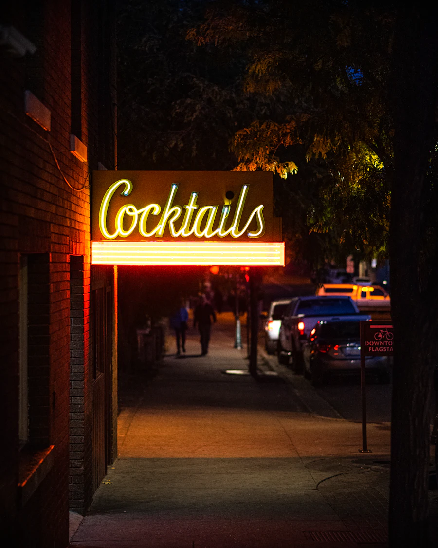 Glowing “Cocktails” neon sign mounted above a dimly lit sidewalk at night, with parked cars and tree shadows receding into the background.