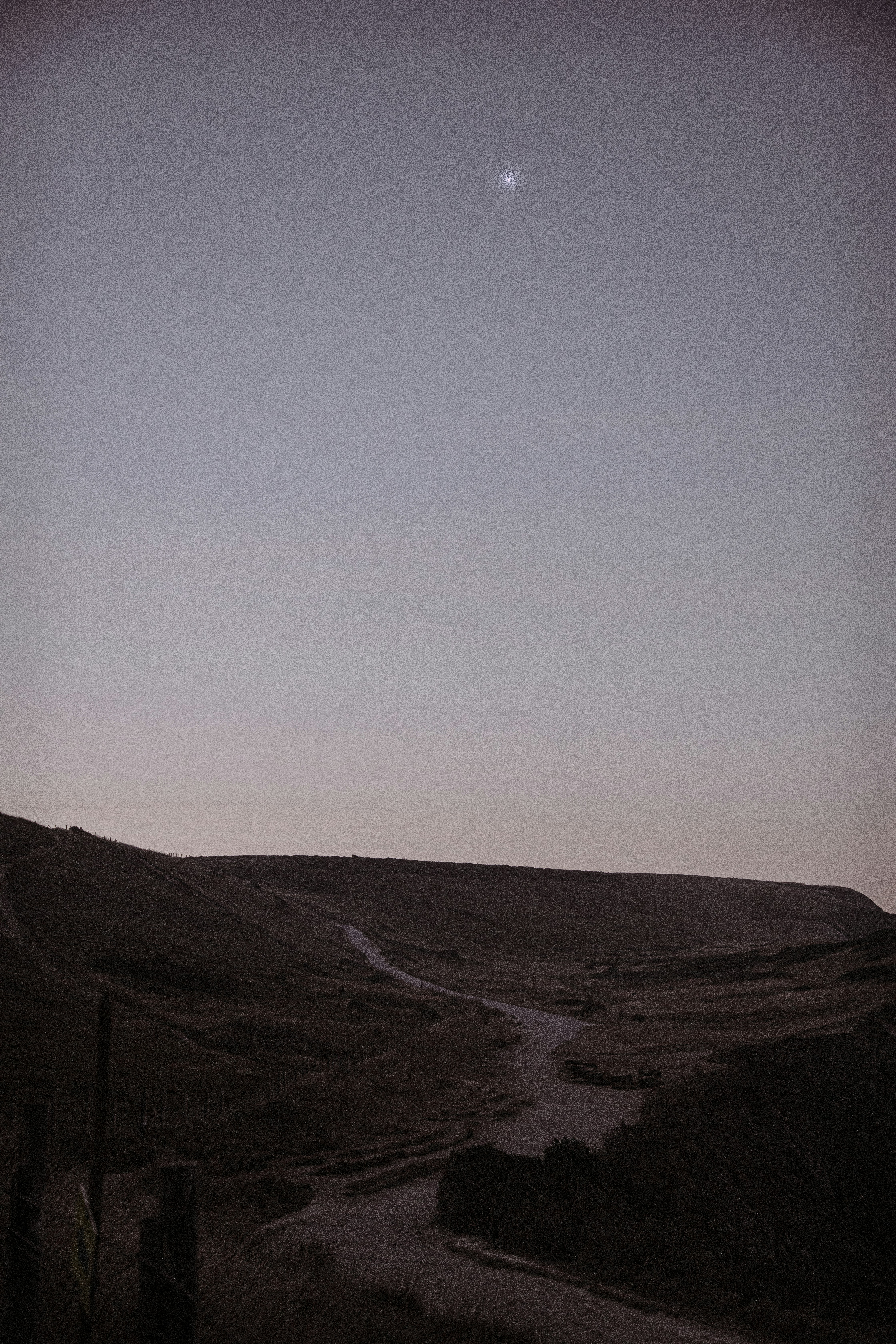 A serene landscape at dusk featuring a winding path through rolling hills under a softening sky. The faint glow of a distant celestial body adds a touch of mystique.
