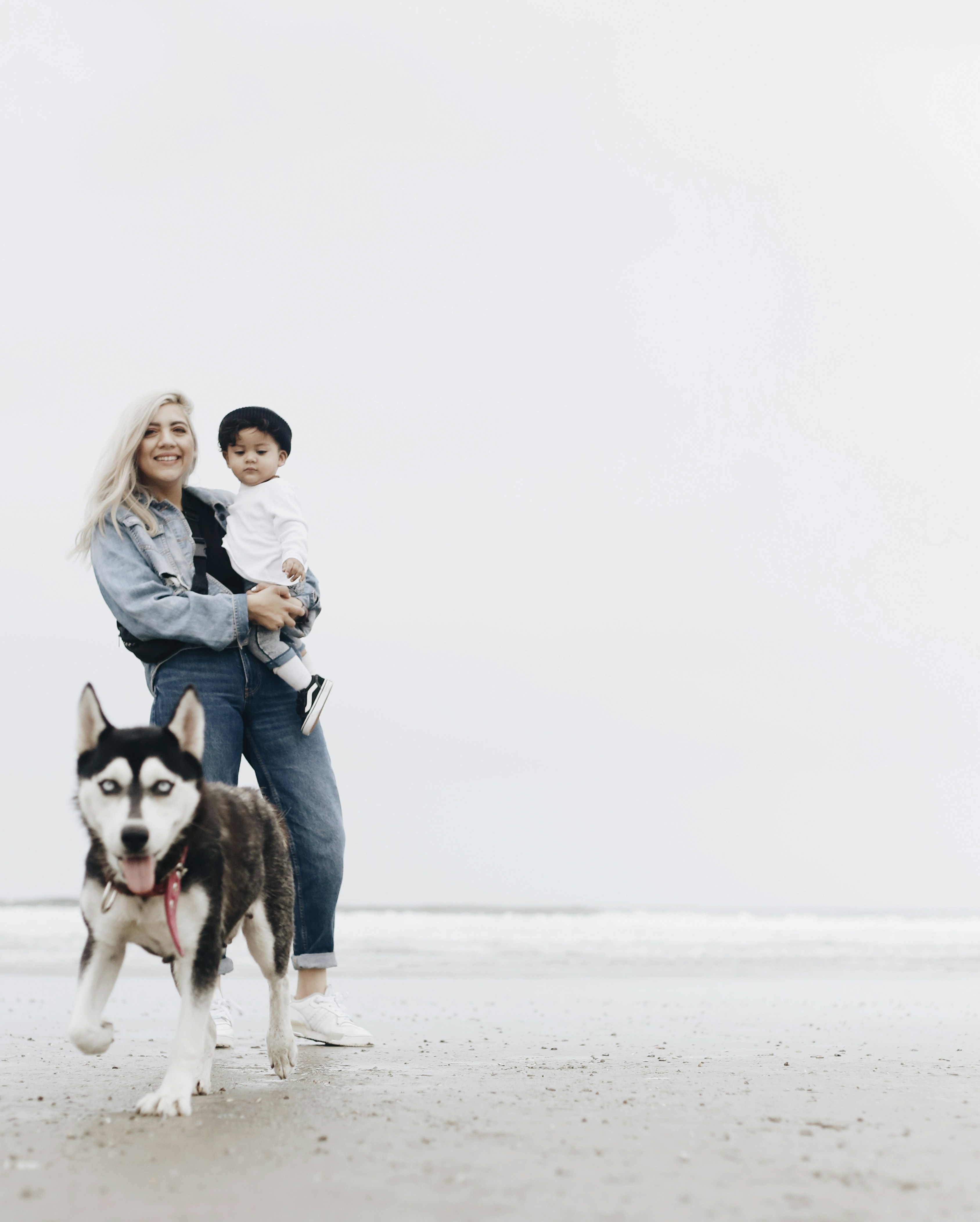 A woman holds a child while a playful husky runs along the beach, capturing a moment of family joy against a serene backdrop.