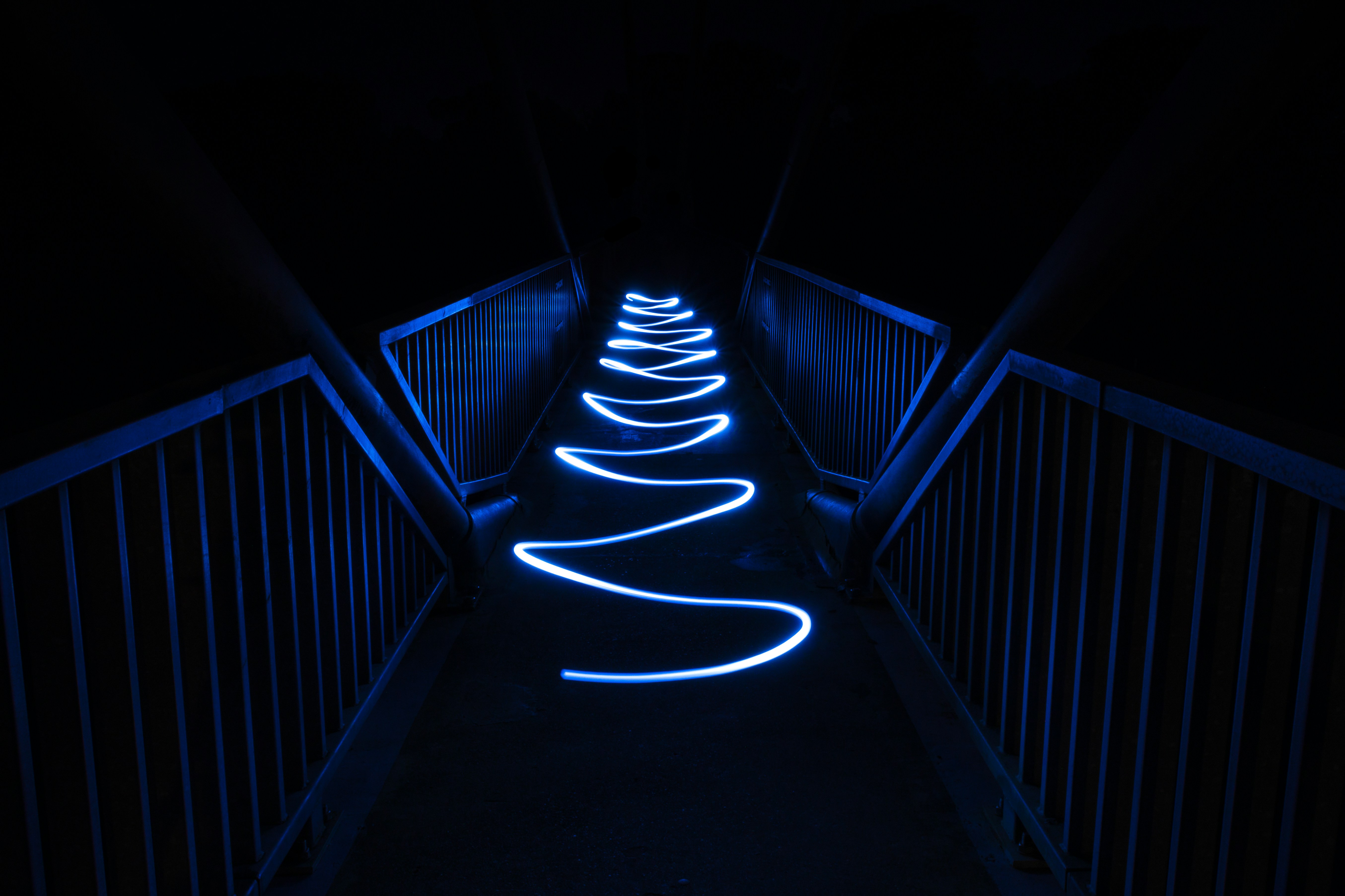 Curving light trails illuminate a dark footbridge at night.