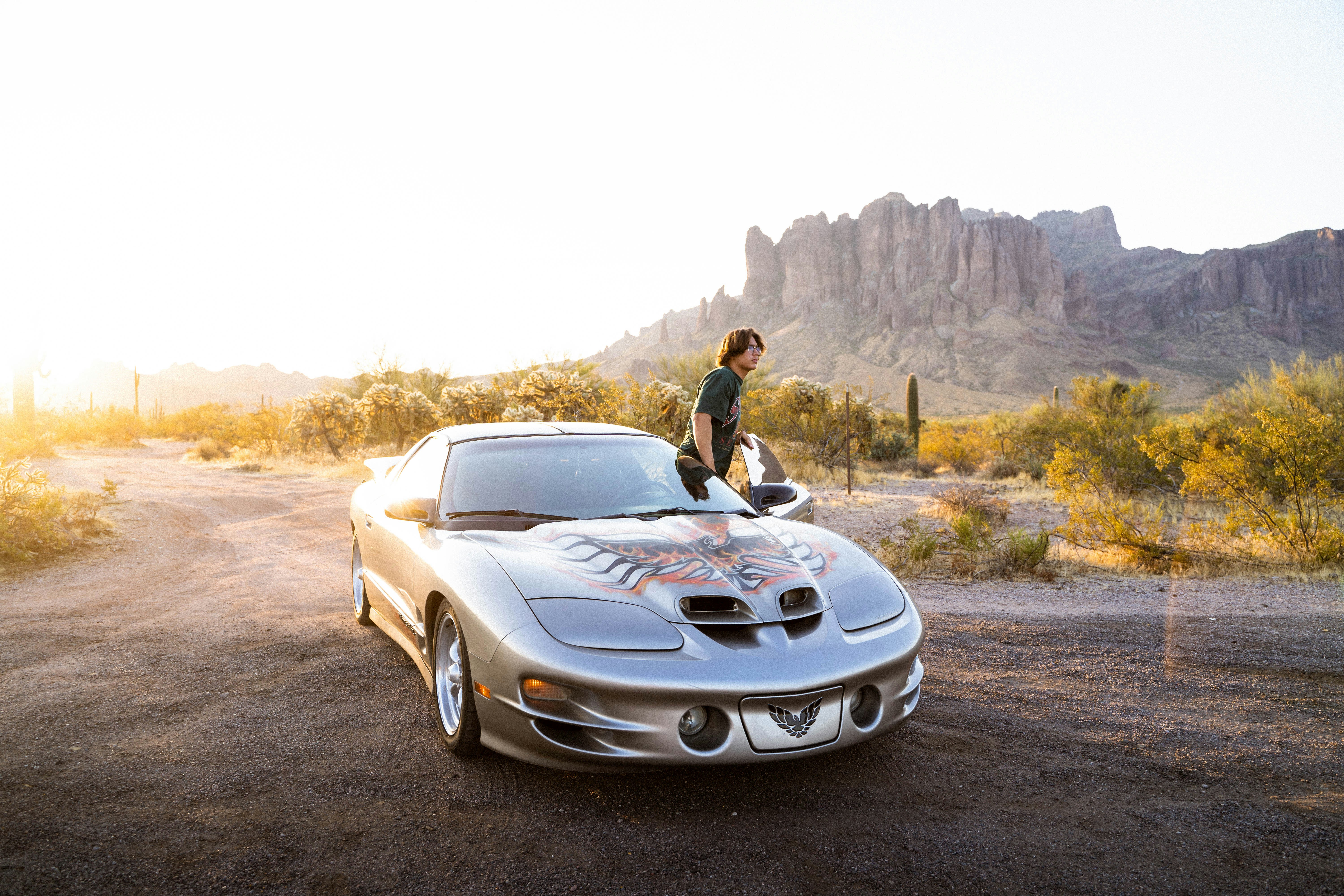 A modified sports car parked on a desert road with a person standing beside it, illuminated by the warm glow of the setting sun.