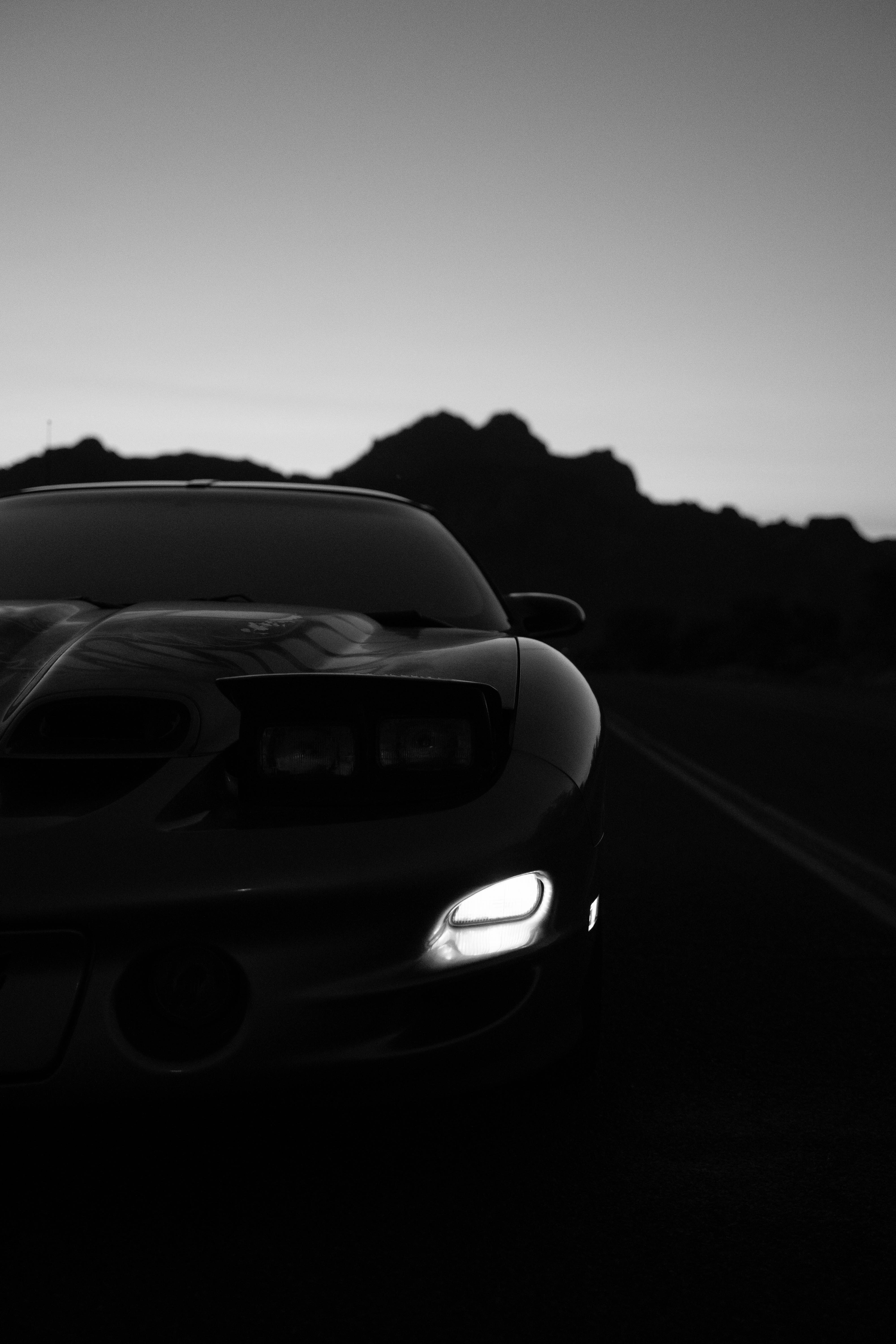 Close-up of a sleek car's front end against a twilight mountain backdrop, emphasizing its headlights and contours.