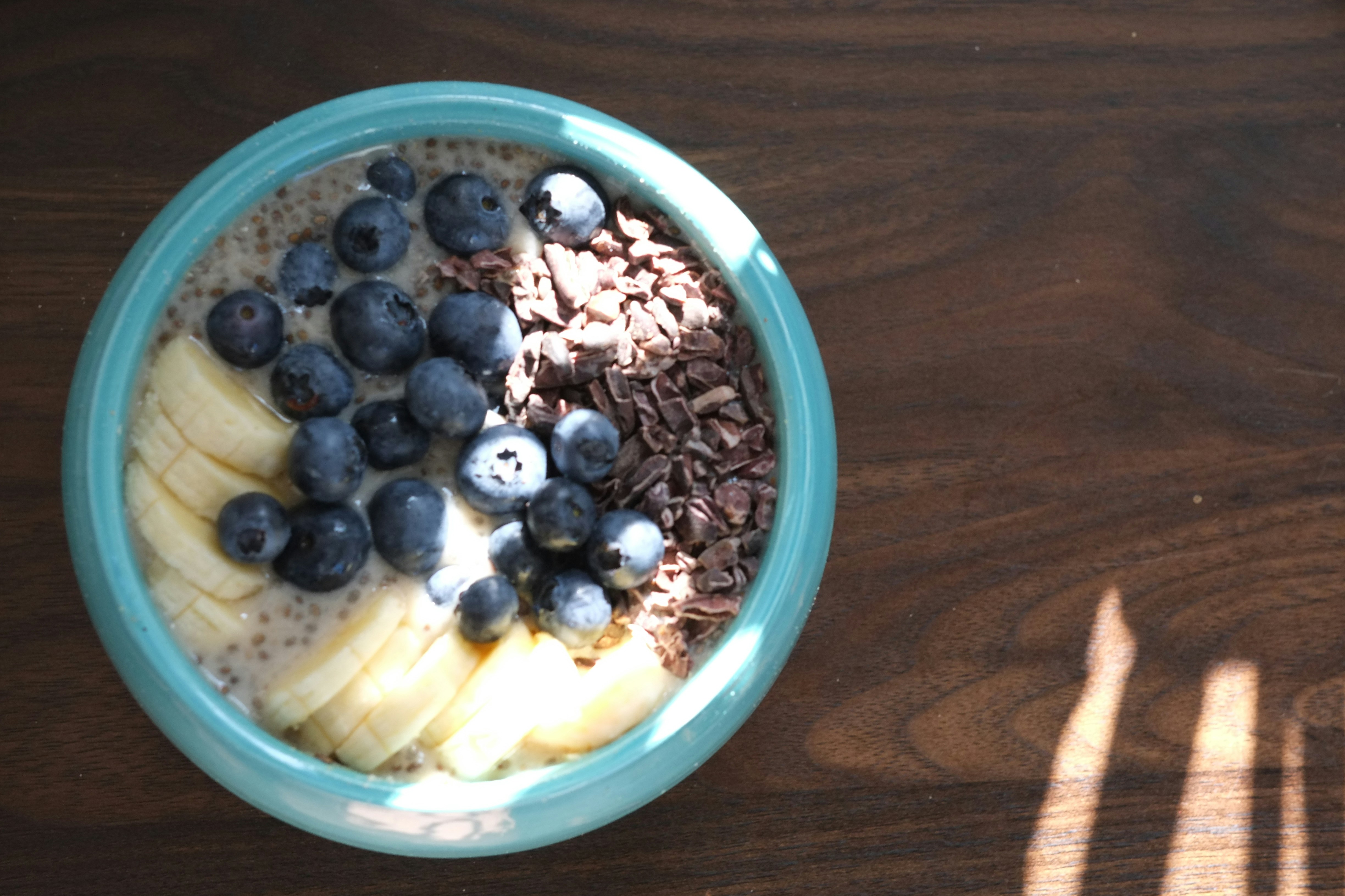 Chia pudding topped with sliced bananas, blueberries, and cacao nibs in a turquoise bowl on a wooden table.