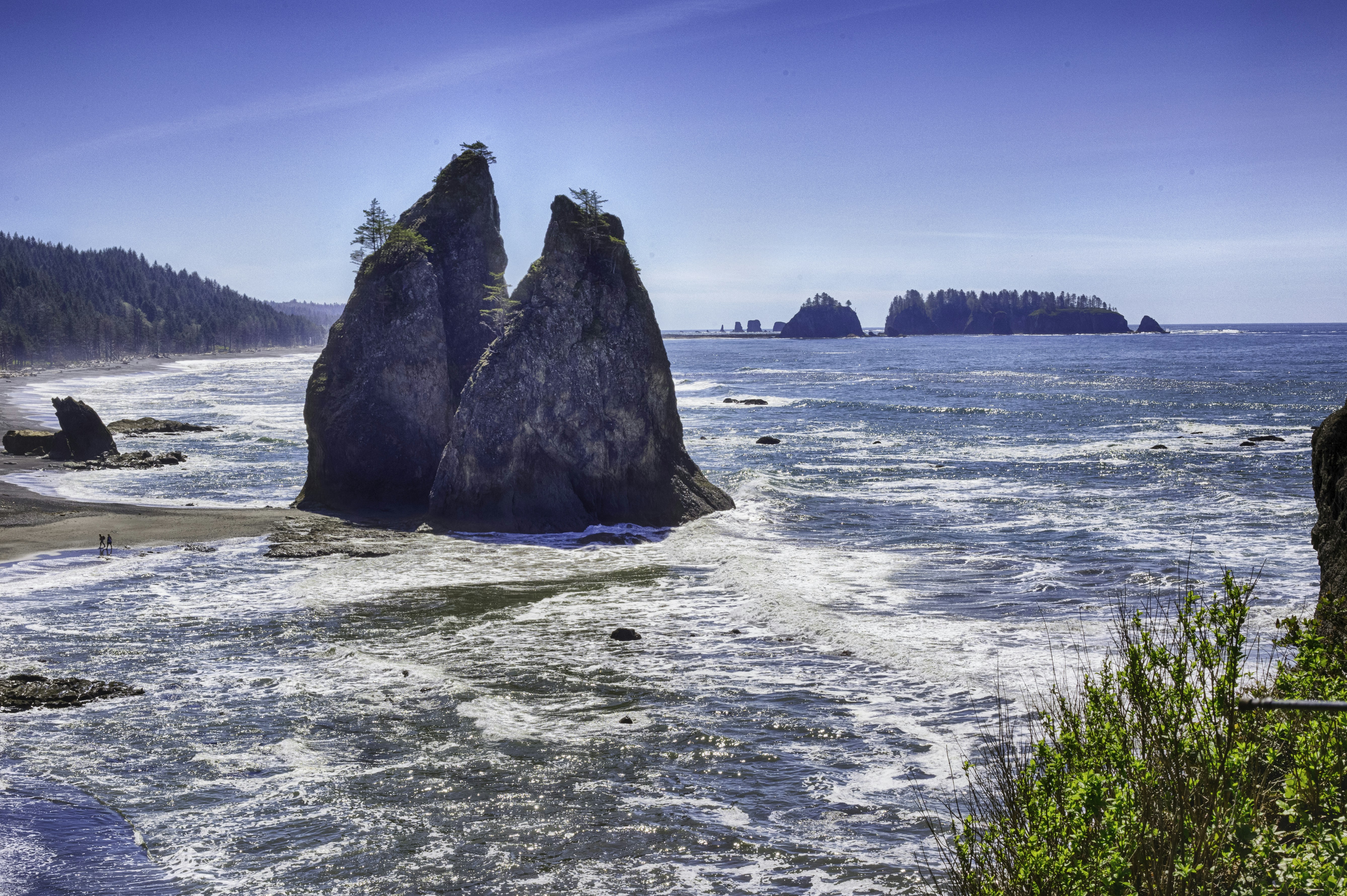 Towering rock formations rise from the ocean near a sandy shore under a clear blue sky.