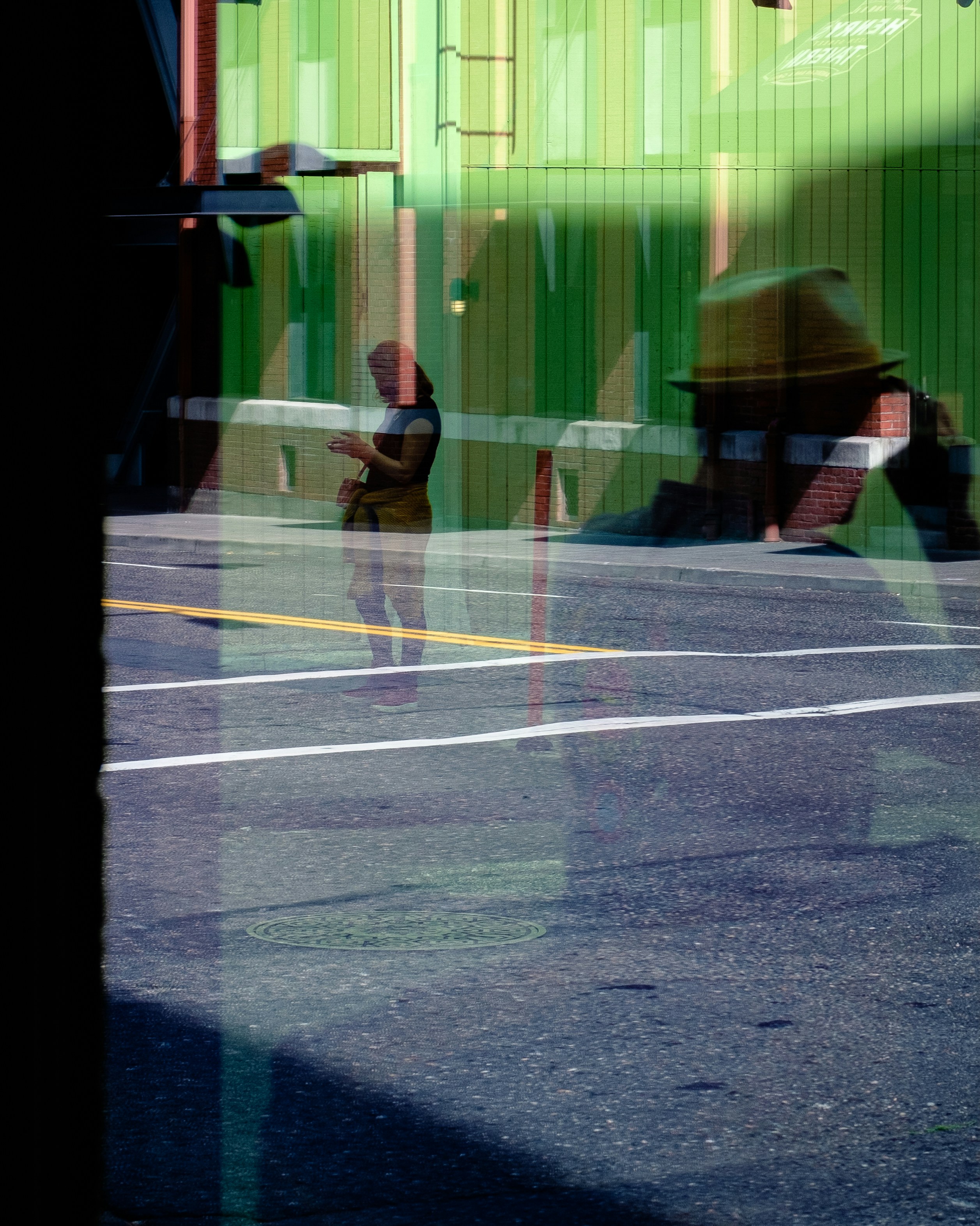 A woman in a yellow top and red headscarf is seen walking on a city street, reflected in a glass surface, with vibrant green building hues in the background.