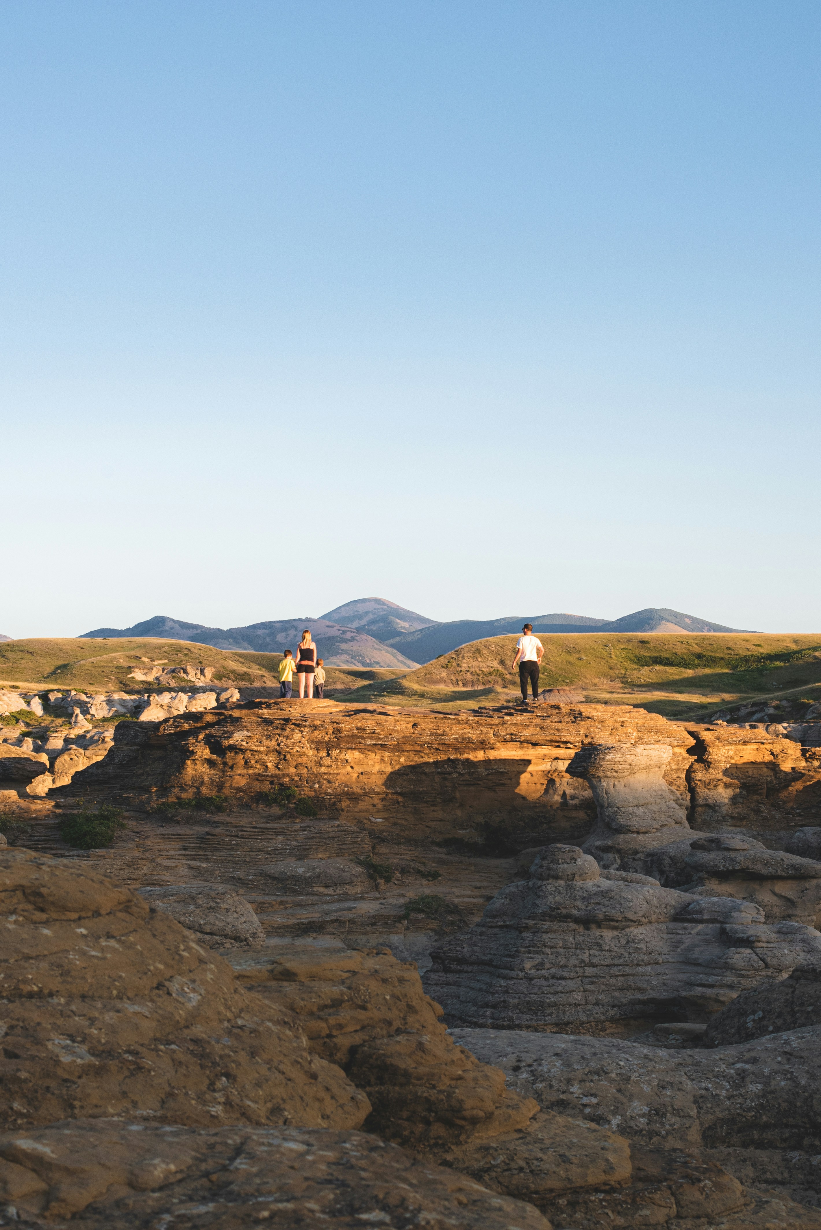 Three individuals standing on a rugged rock formation, overlooking rolling hills and distant mountains under a clear sky.