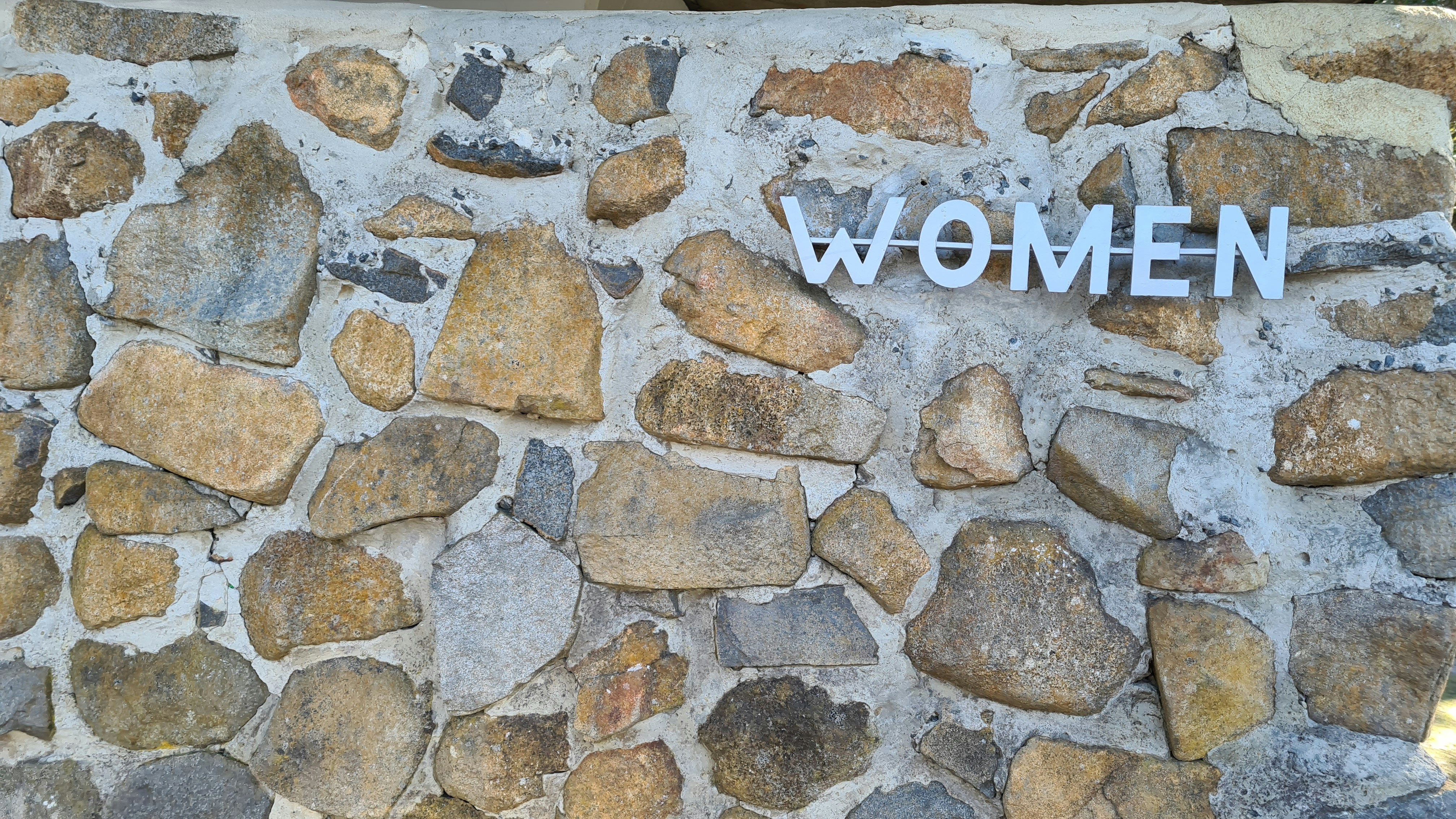 Metal letters spelling 'WOMEN' mounted on a rustic stone wall. The stark typography contrasts with textured masonry, emphasizing urban signage.