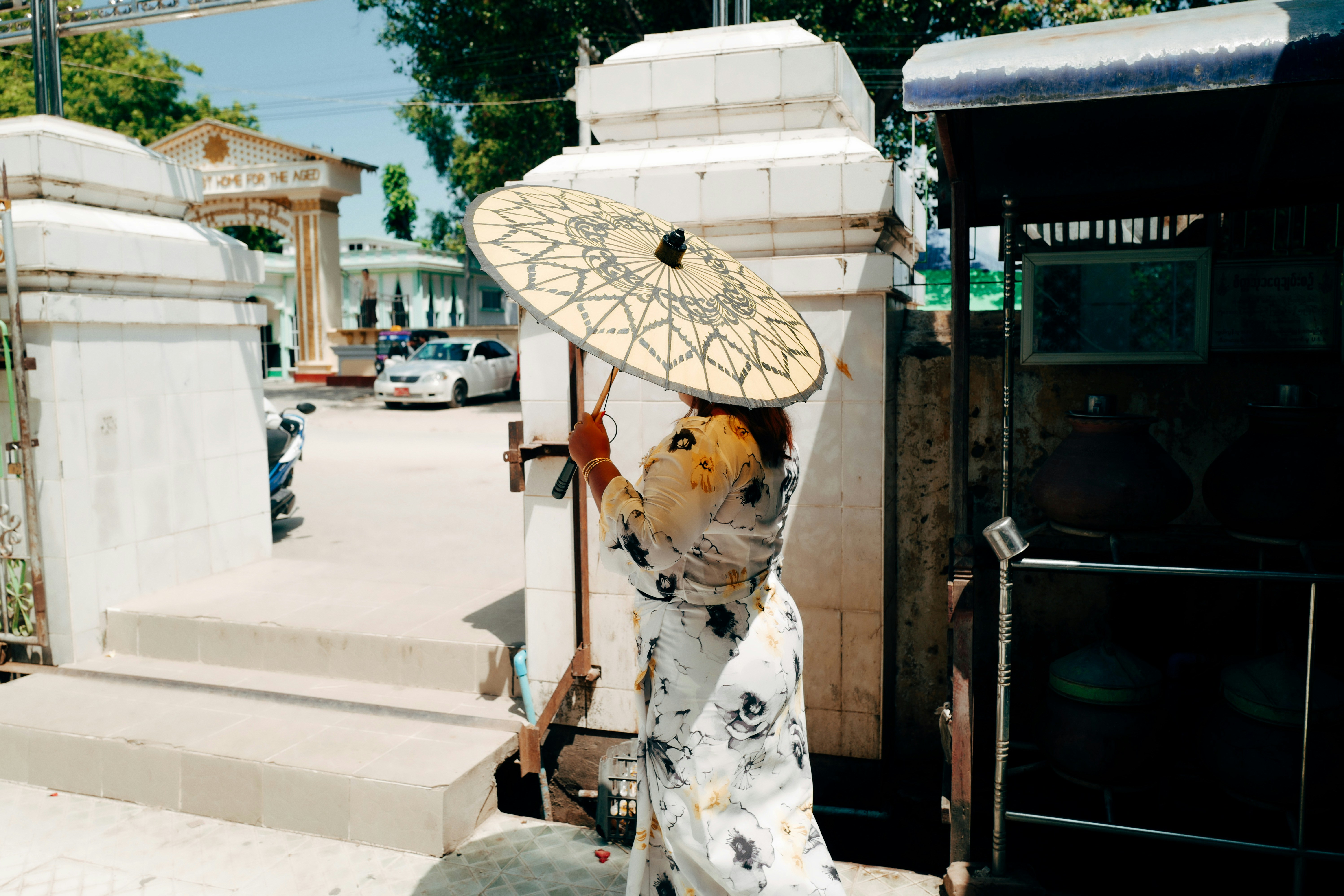 Woman in a patterned kimono holding a decorative parasol, walking through a vibrant outdoor scene. The setting features a blend of traditional architecture and modern elements.