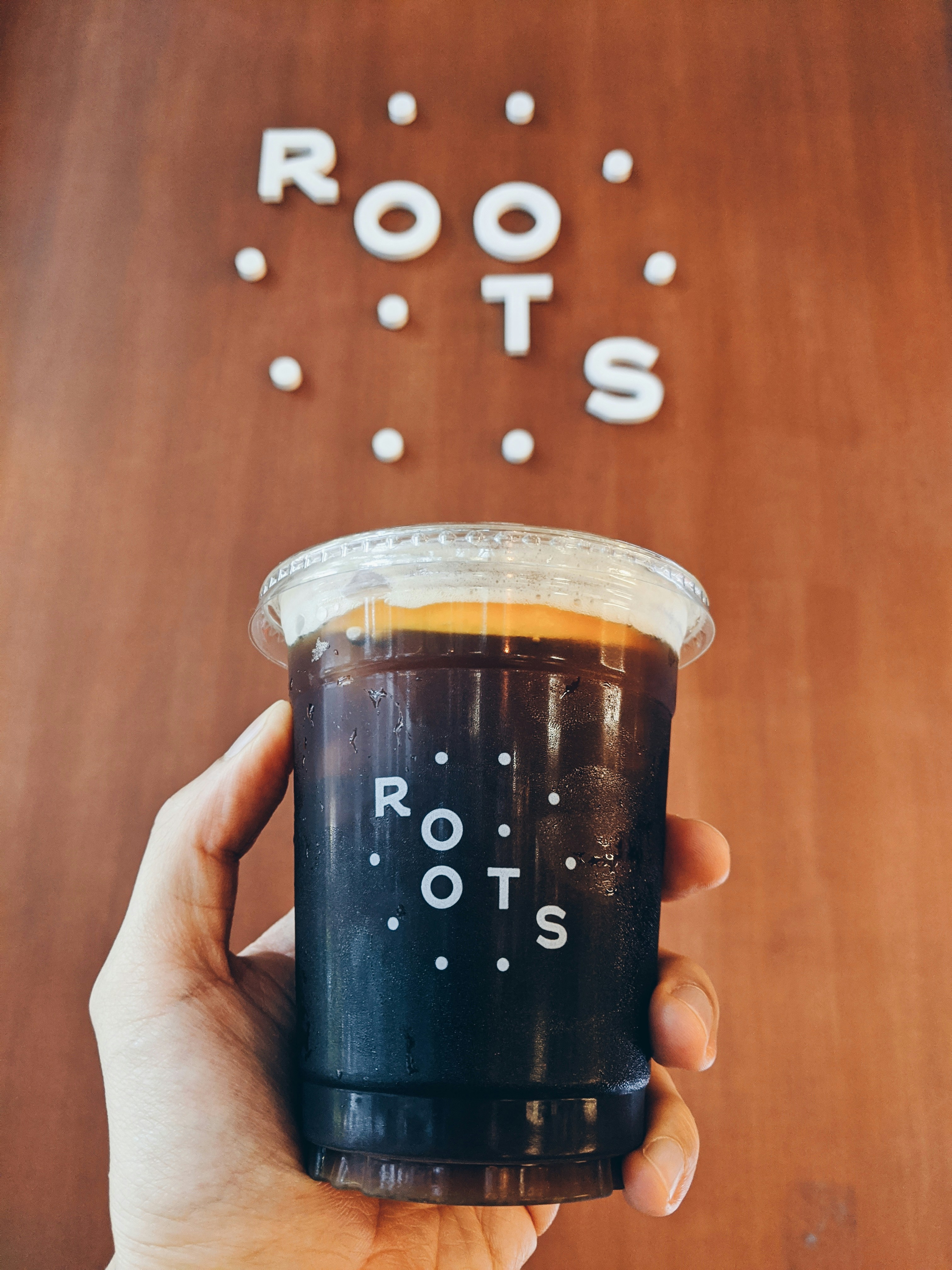 Hand holding a cup of iced coffee with the logo 'ROOTS' prominently displayed, set against a backdrop of playful letter arrangements.