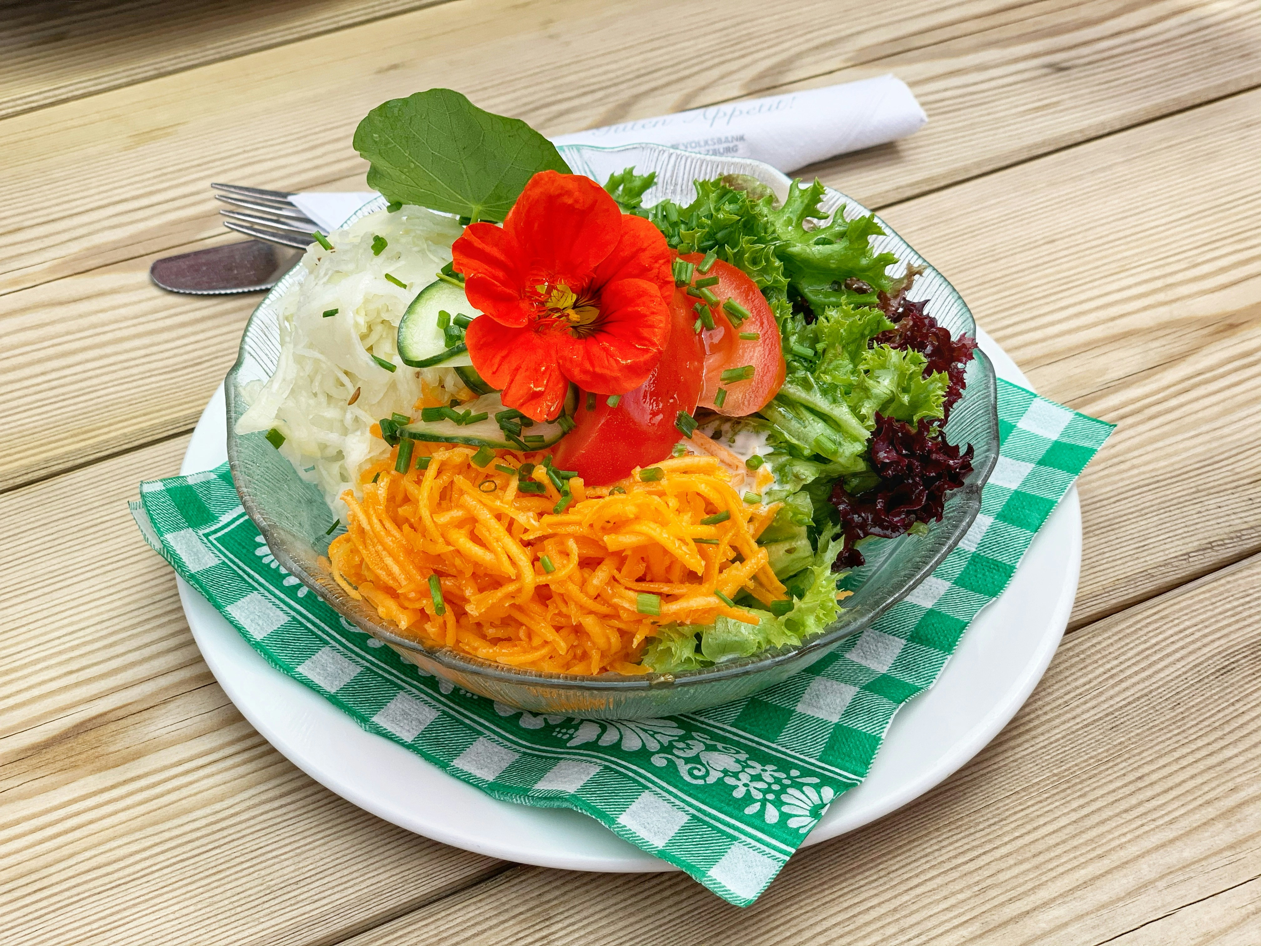 Colorful salad featuring a mix of greens, shredded carrots, tomato slices, and an edible flower, elegantly presented on a checkered napkin.