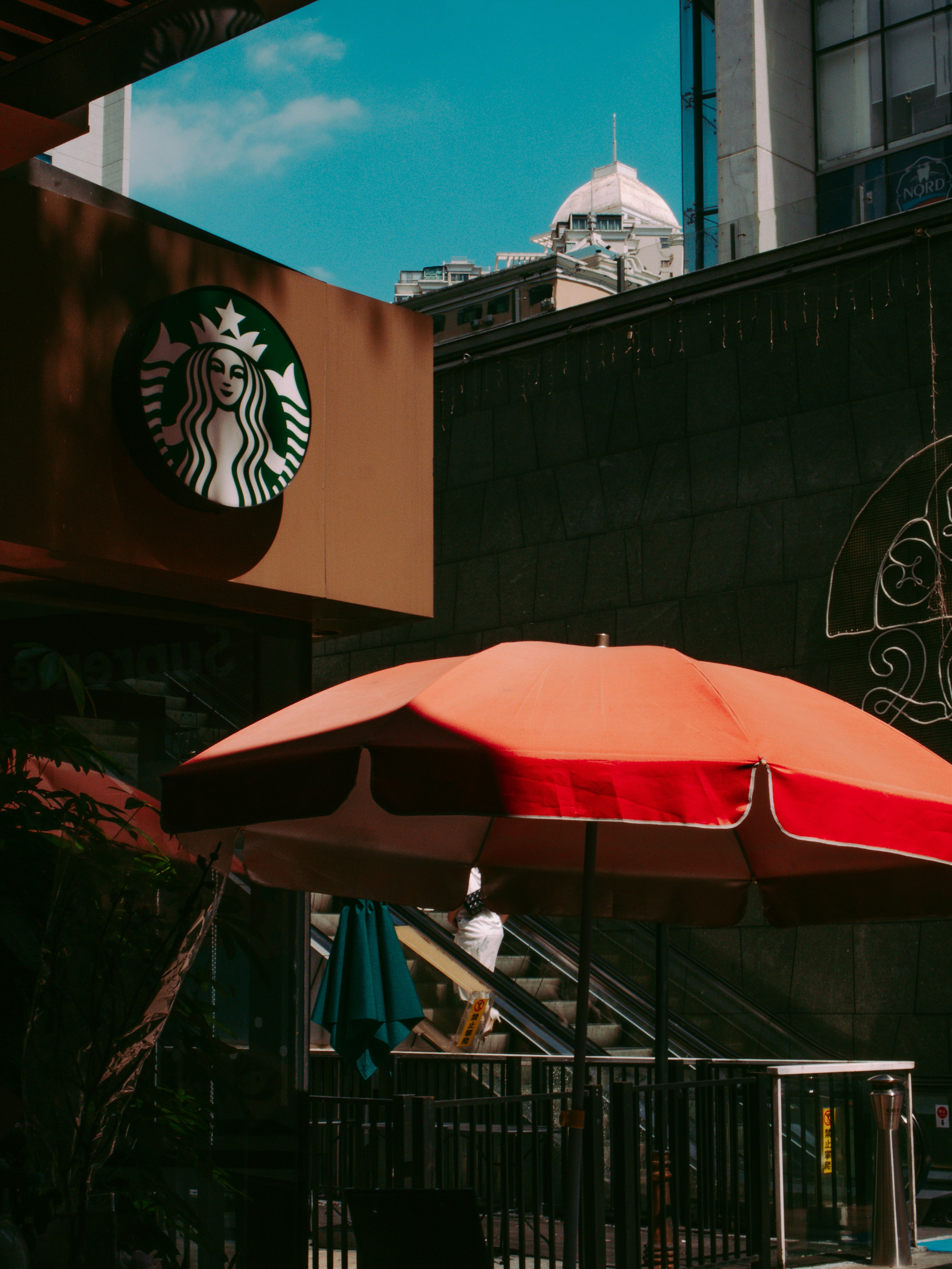 Bright red umbrella providing shade in a bustling urban setting near a Starbucks logo, with a modern architectural backdrop.
