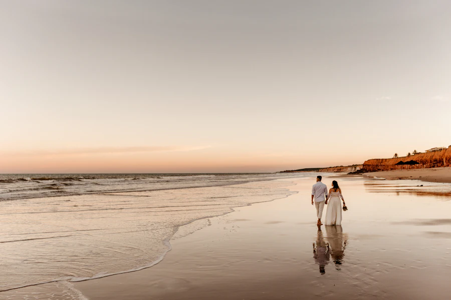 A couple in light clothing walks hand in hand along a reflective shoreline at sunset, with gentle waves and a warm sky behind them. Cliffs and beach huts line the distant right edge.