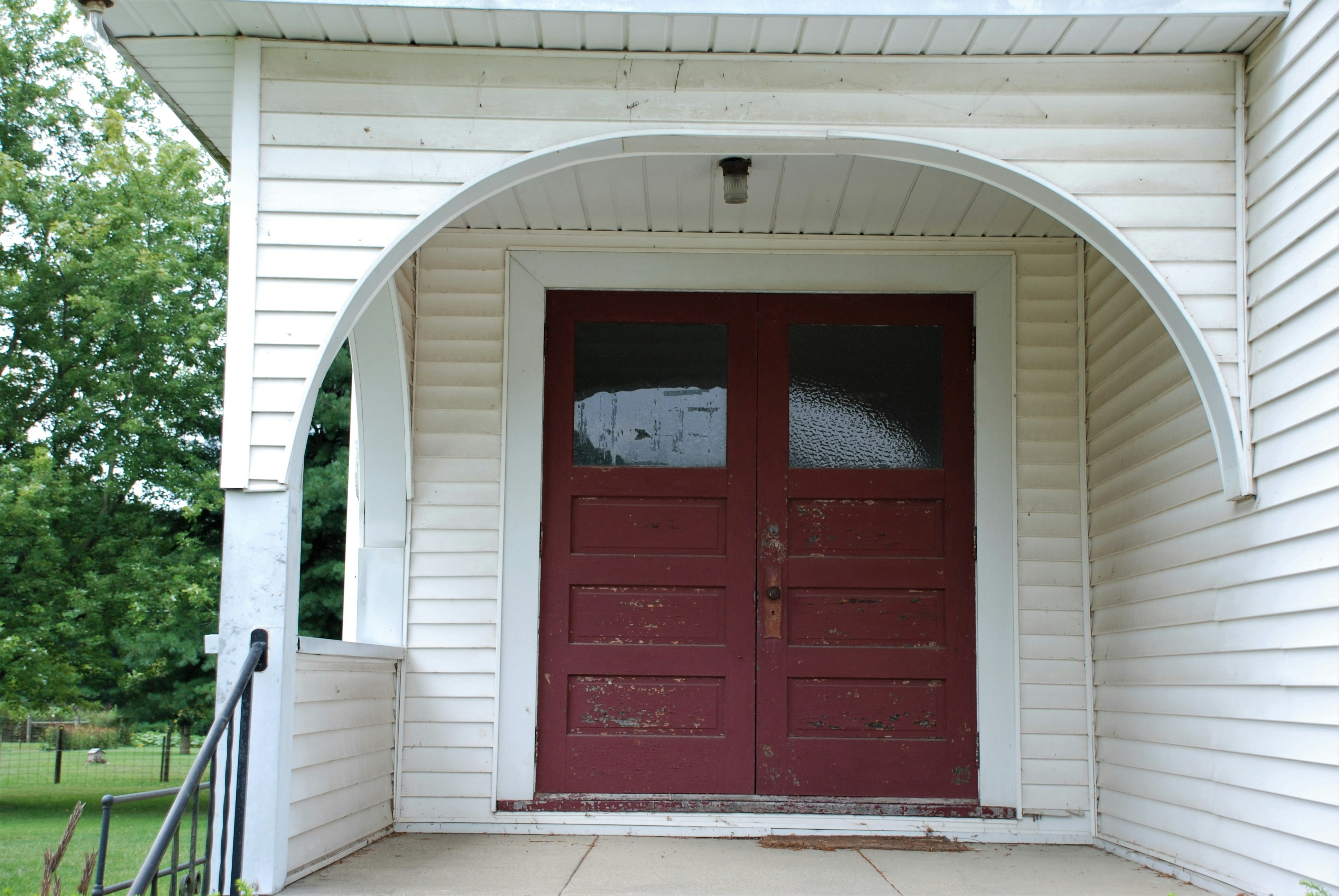 A pair of vintage red doors with peeling paint, framed by a white archway, set against a backdrop of lush greenery.