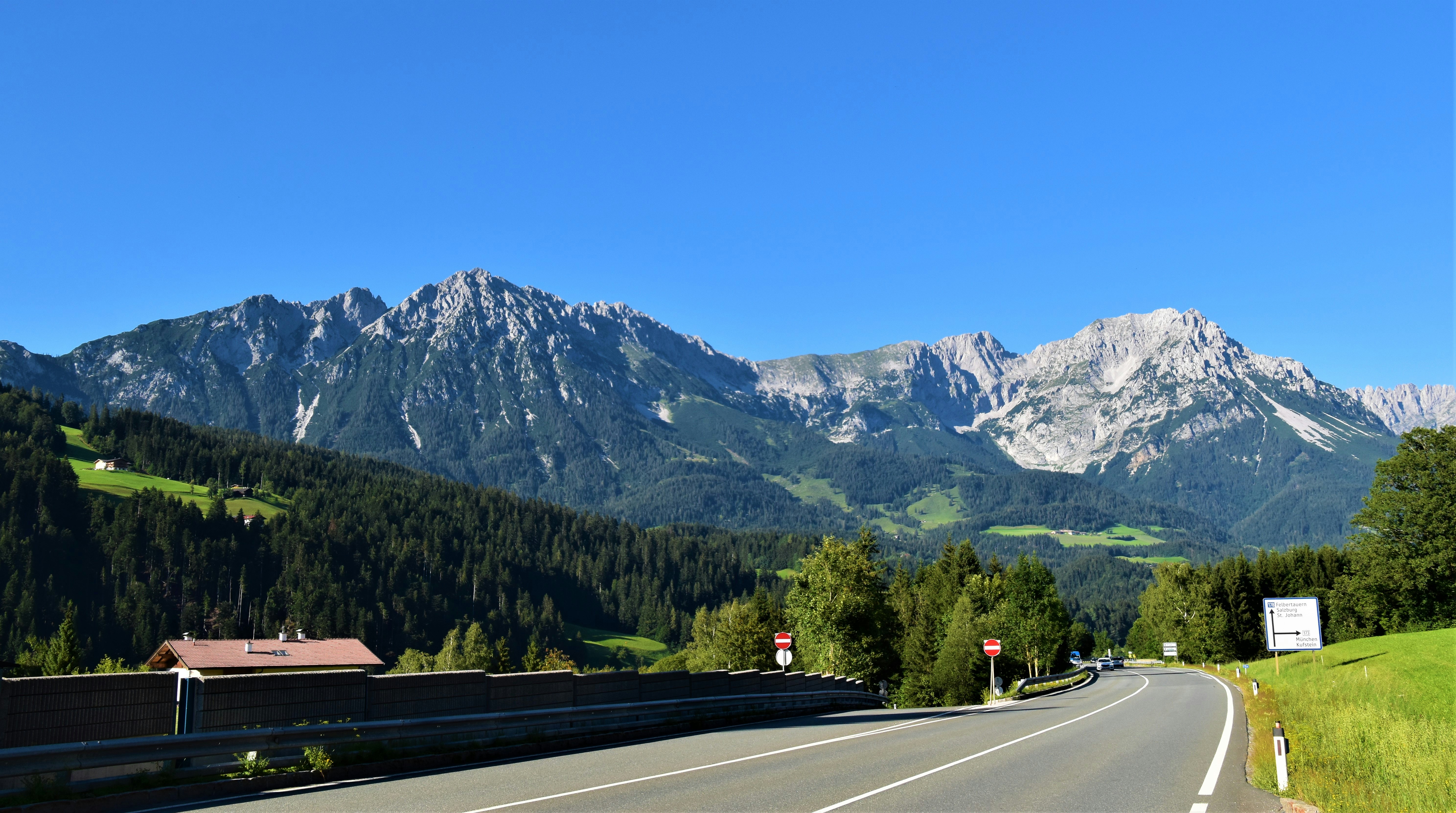 Winding road leading towards the impressive alpine mountains under a clear blue sky. Lush greenery flanks the highway, enhancing the scenic beauty.