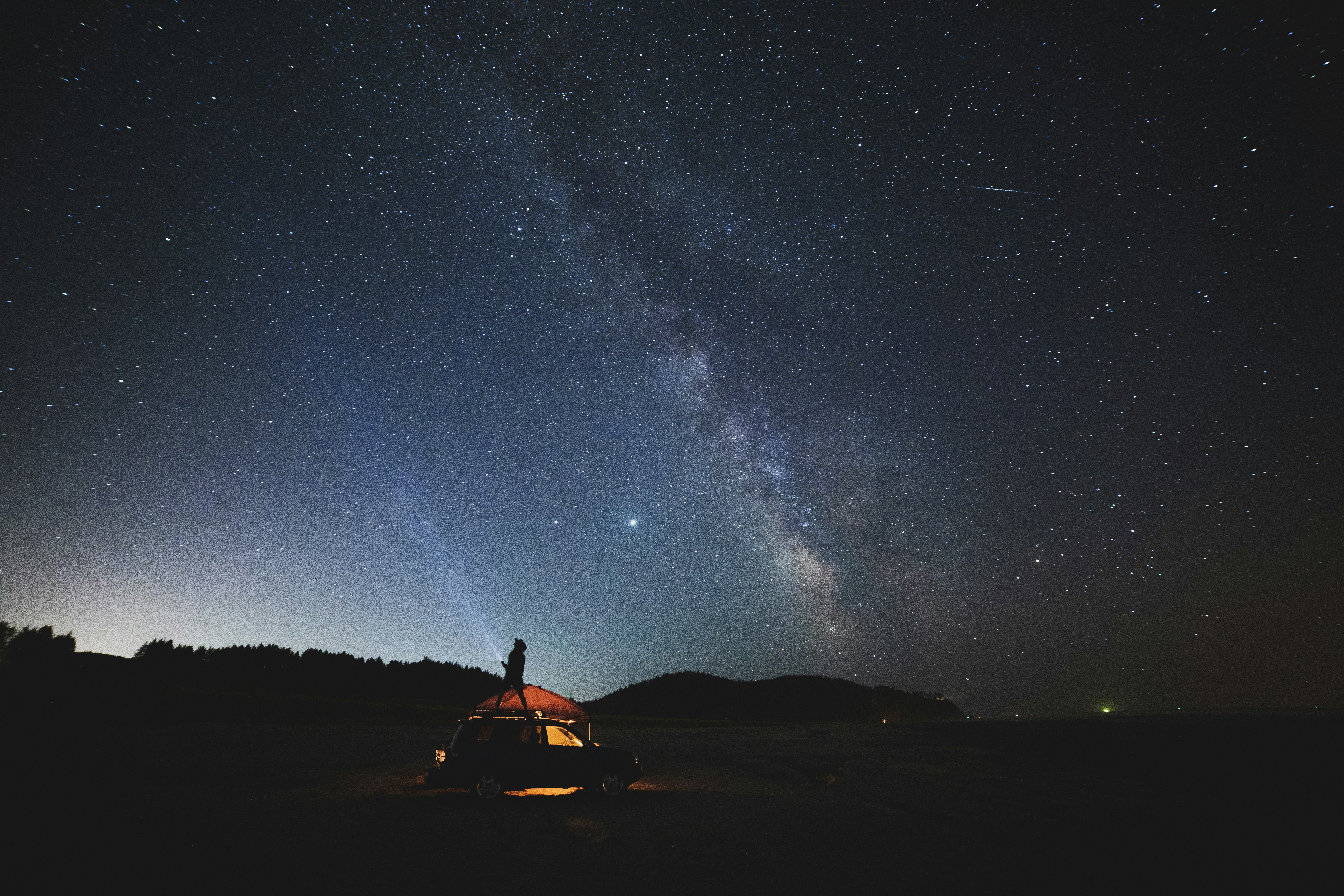 A figure stands atop a car, gazing at the expansive Milky Way in a star-filled sky. The scene captures the essence of stargazing in nature.