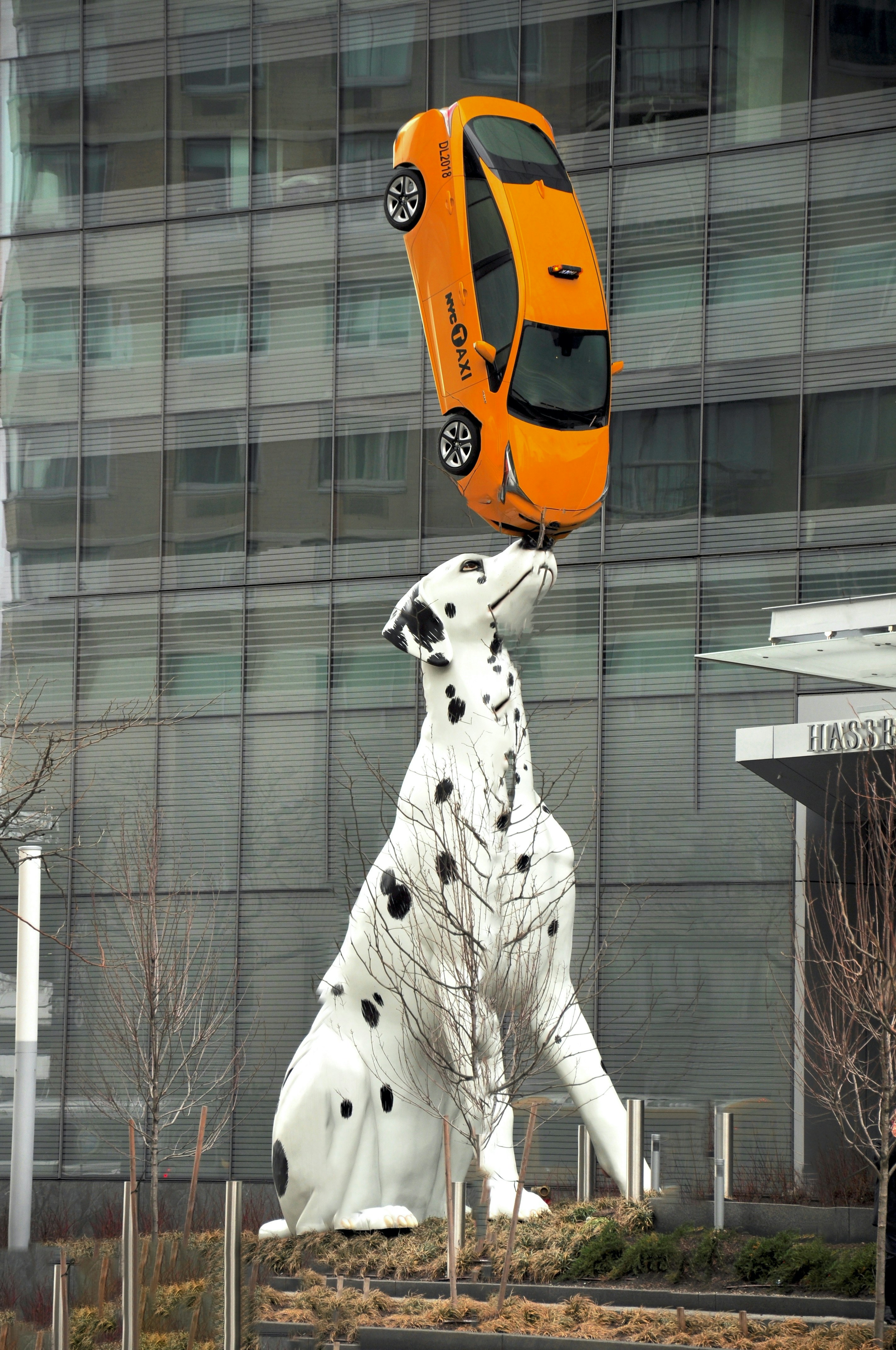 Icarus on a Dalmatian: An orange car perched atop a statue before glass towers