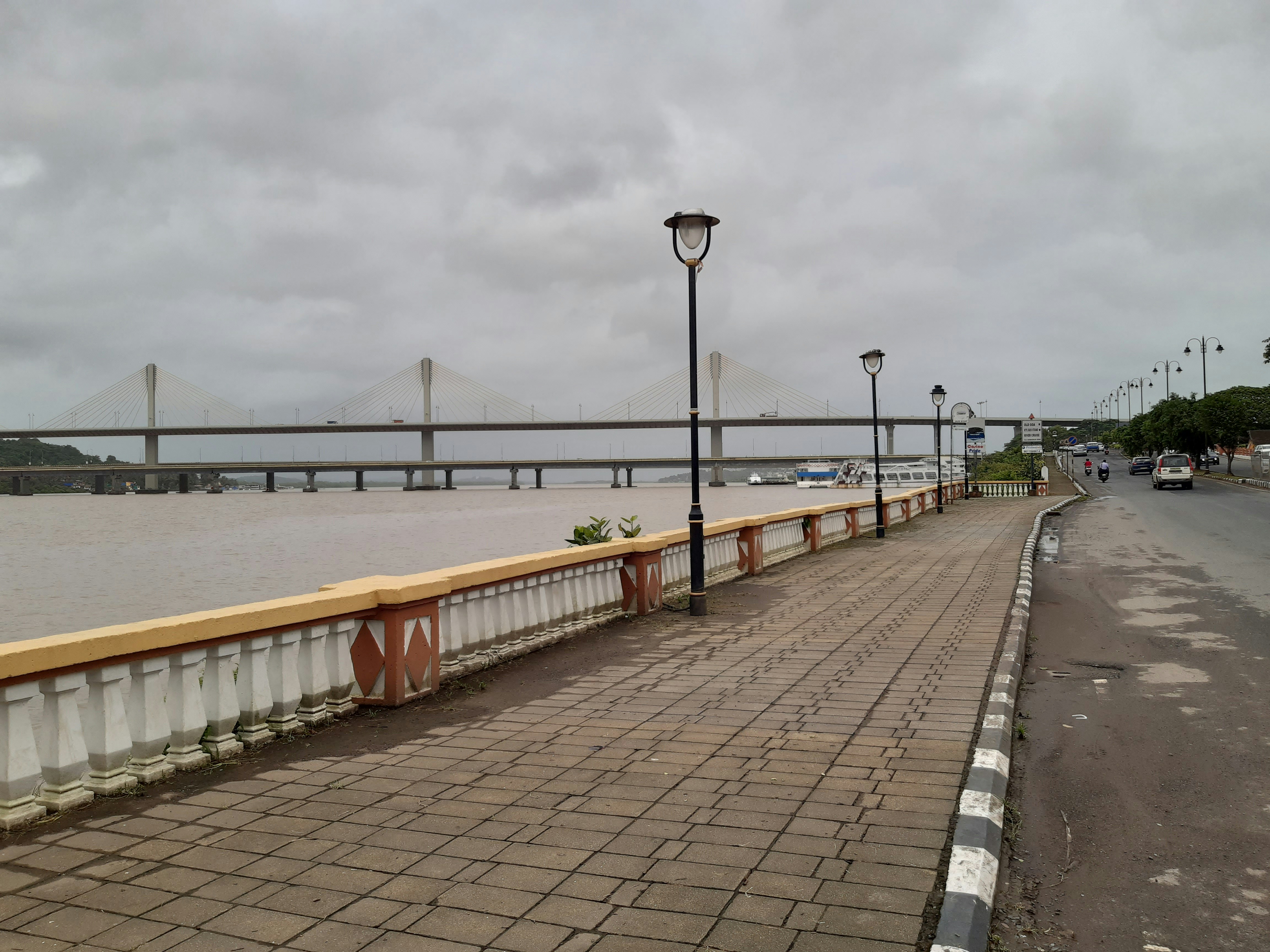 Overcast skies cast a muted light on a paved riverside walkway with a distant view of a large suspension bridge.