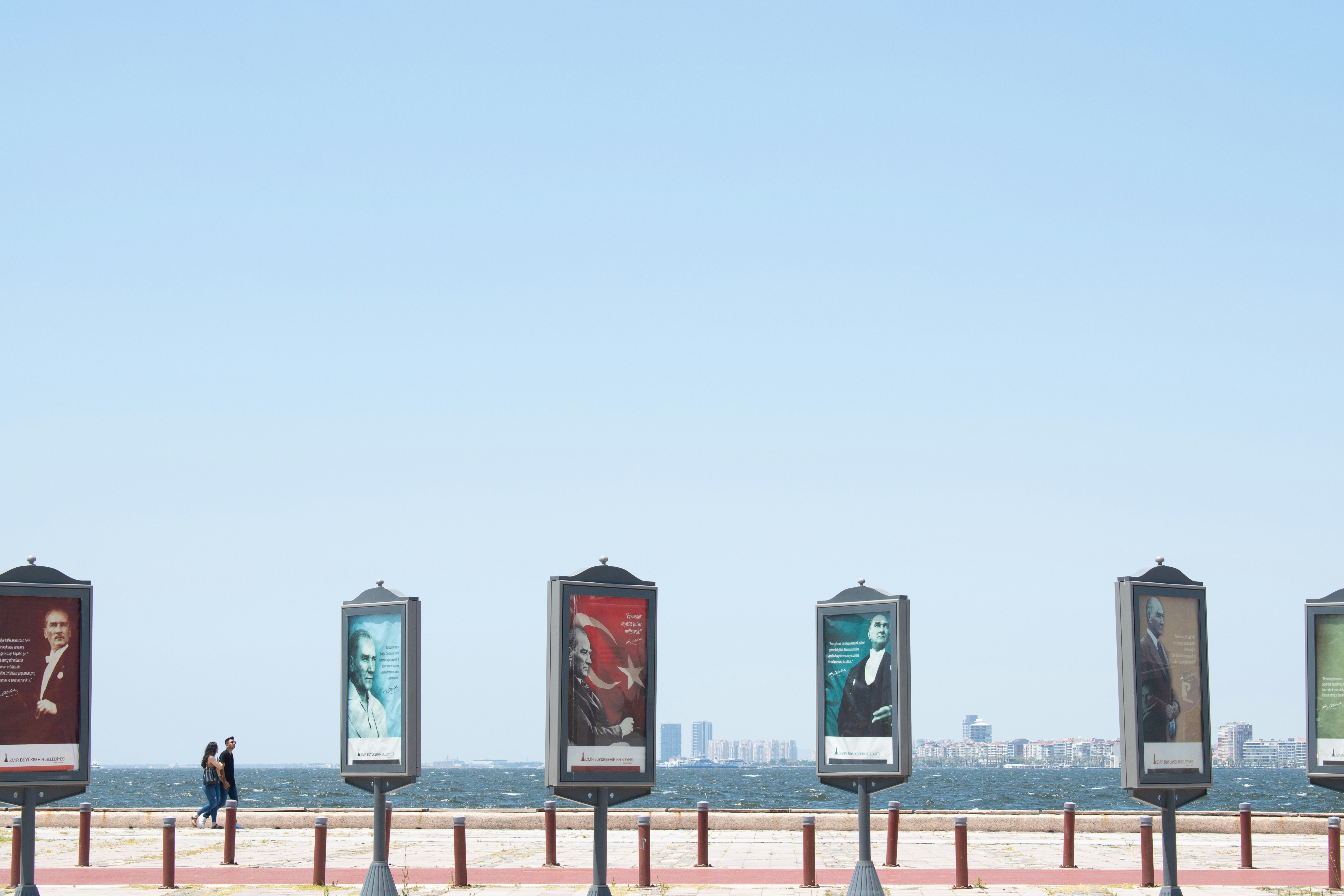 Series of display boards with portraits along a waterfront promenade under a clear blue sky.