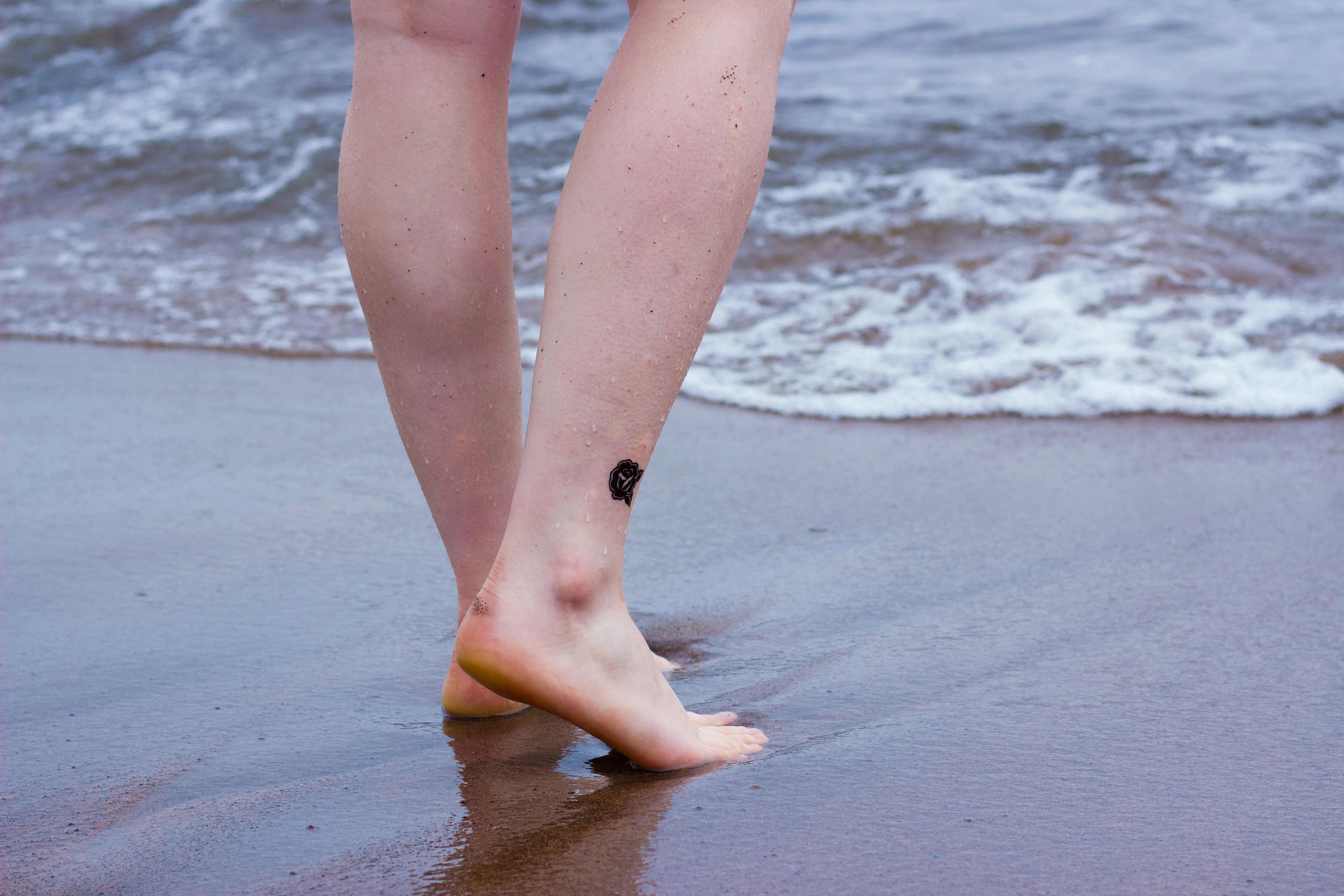 Barefoot legs wade through gentle ocean waves, revealing a delicate tattoo on the ankle. The soft texture of wet sand contrasts with the ebbing tide.