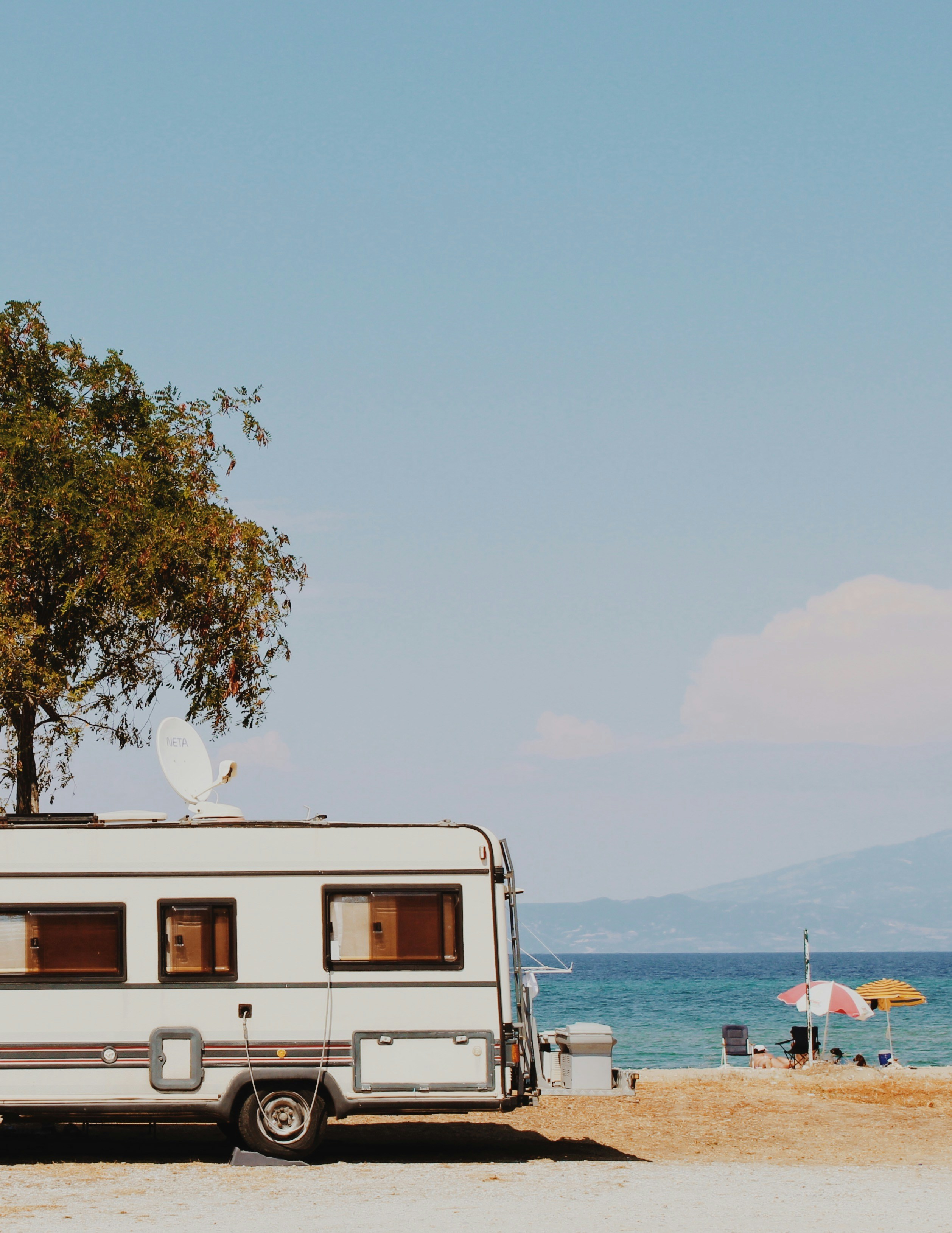 A vintage camper parked under a tree beside a tranquil beach, with colorful umbrellas dotting the sand in the background.