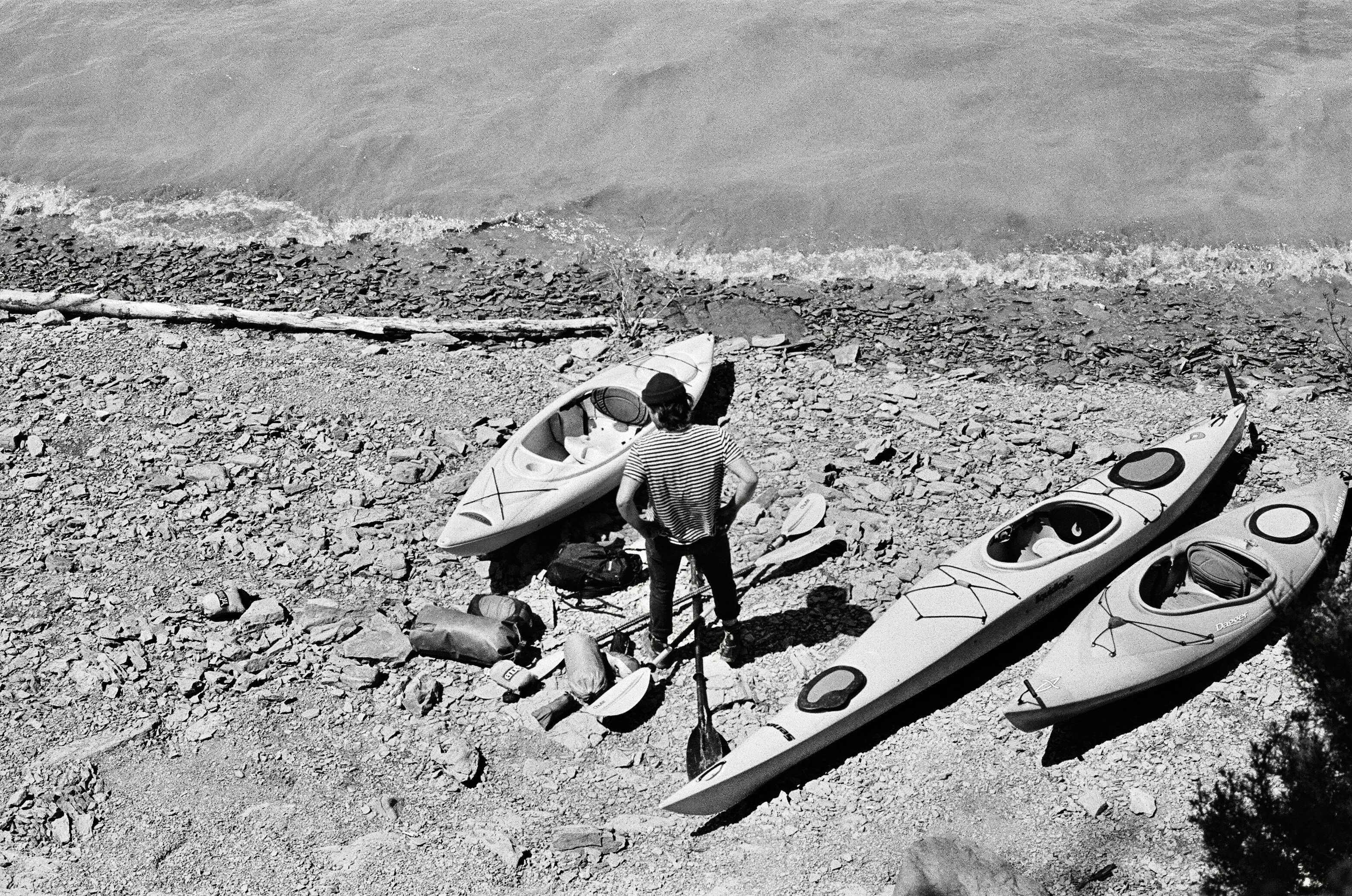 A kayaker stands on a rocky shore, surrounded by kayaks and gear, ready to embark on a water adventure.