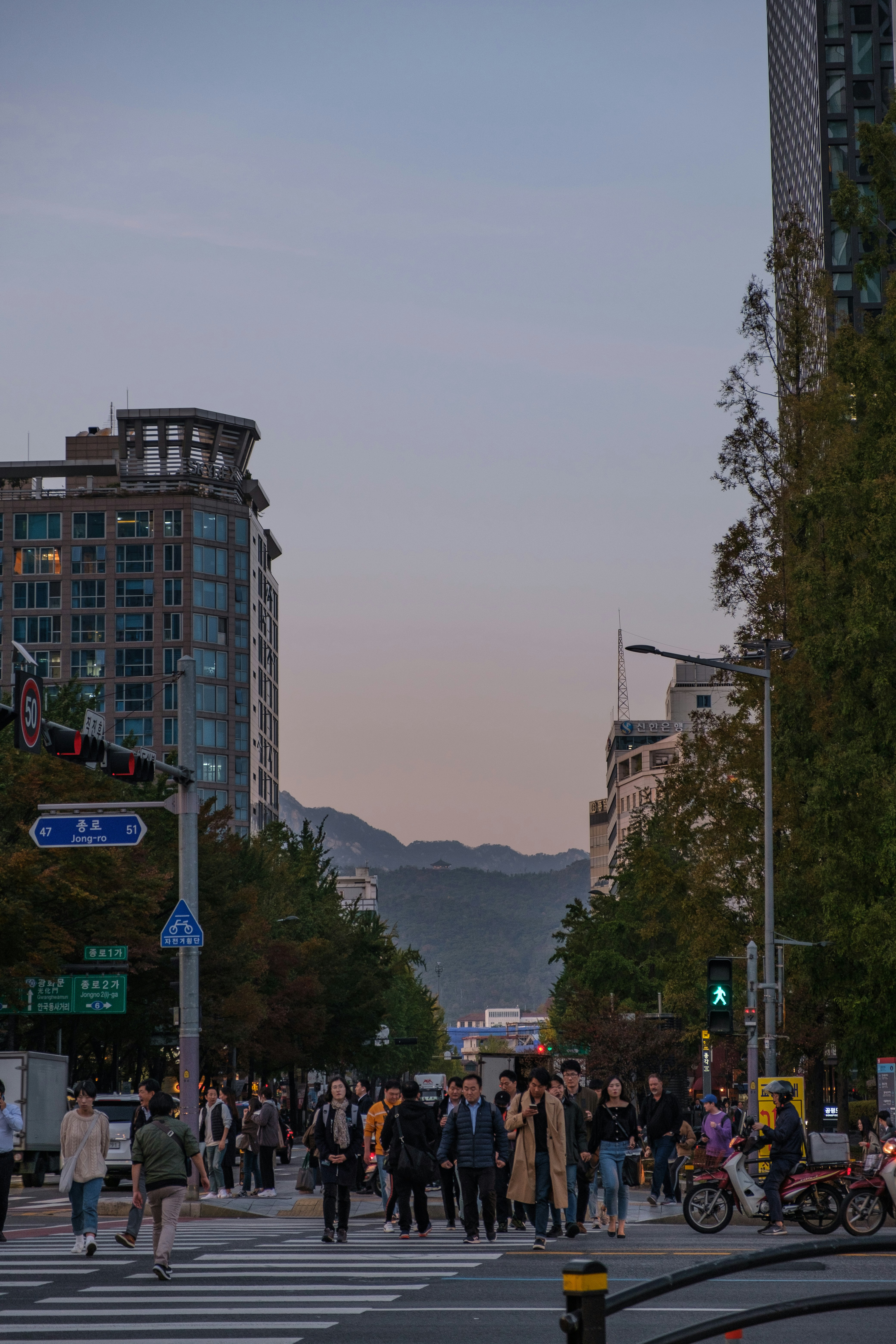 Busy city street bustling with pedestrians crossing at dusk, framed by modern architecture and distant mountains. Traffic signals and street signs add to the urban ambiance.