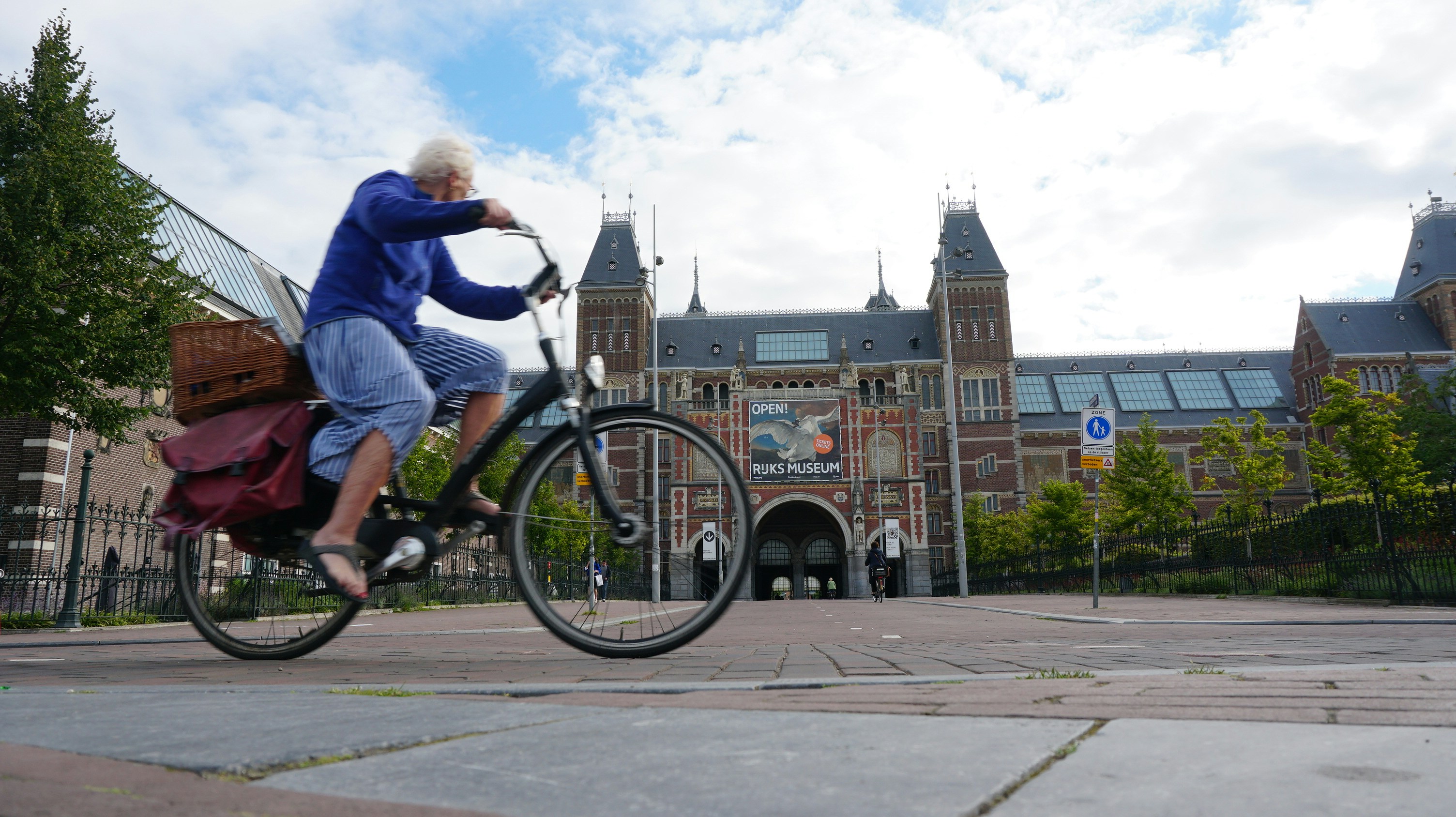 Elderly cyclist navigating the cobblestone path in front of a historic museum, framed by lush greenery and a dramatic sky.