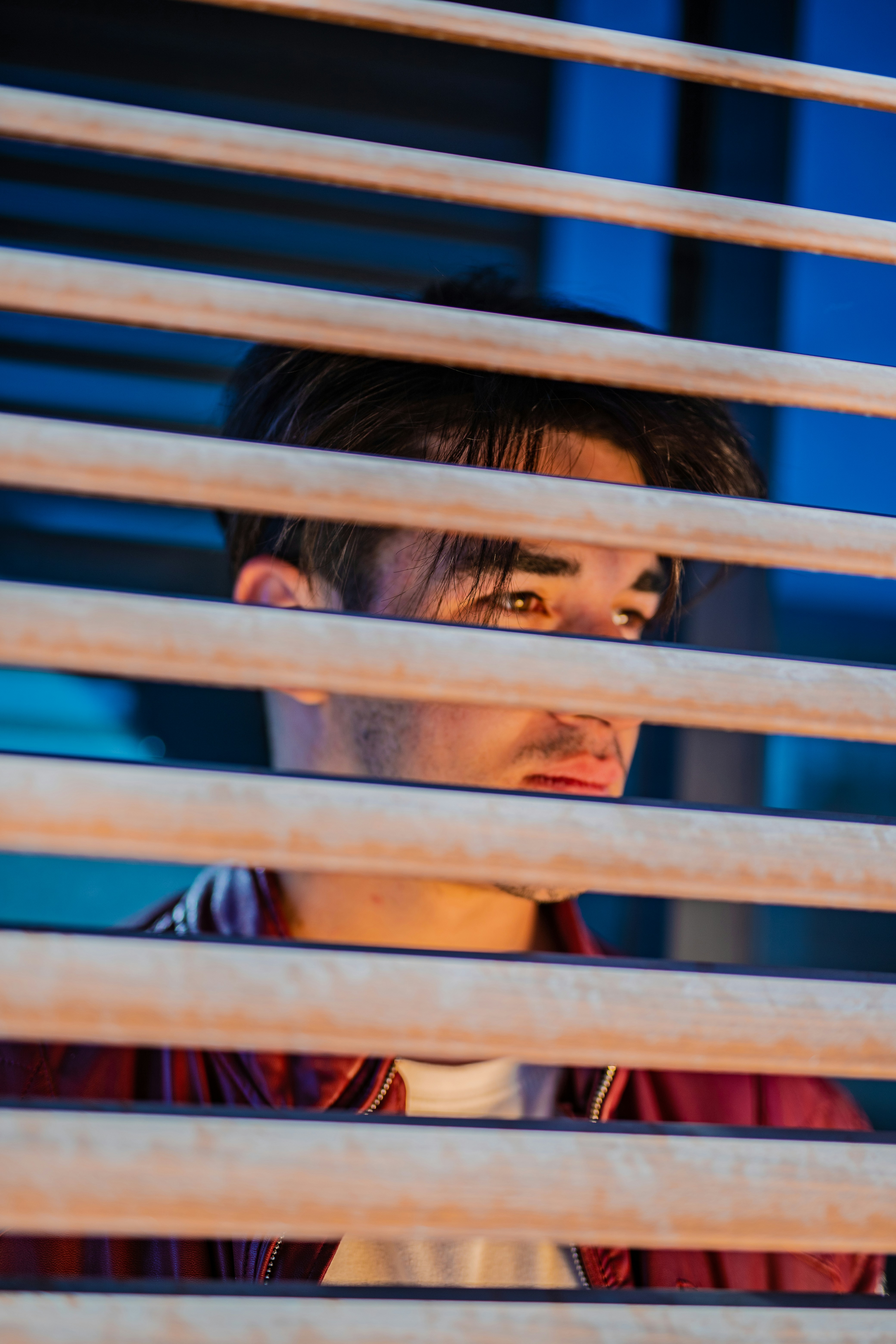 Young man peers through horizontal blinds with a contemplative expression.