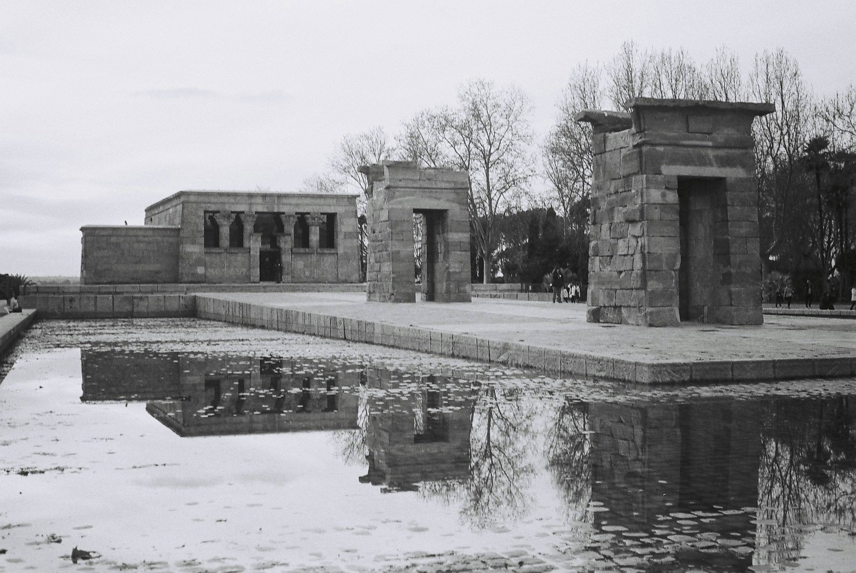 Ancient Egyptian temple ruins reflected in still water, surrounded by bare trees under a cloudy sky.