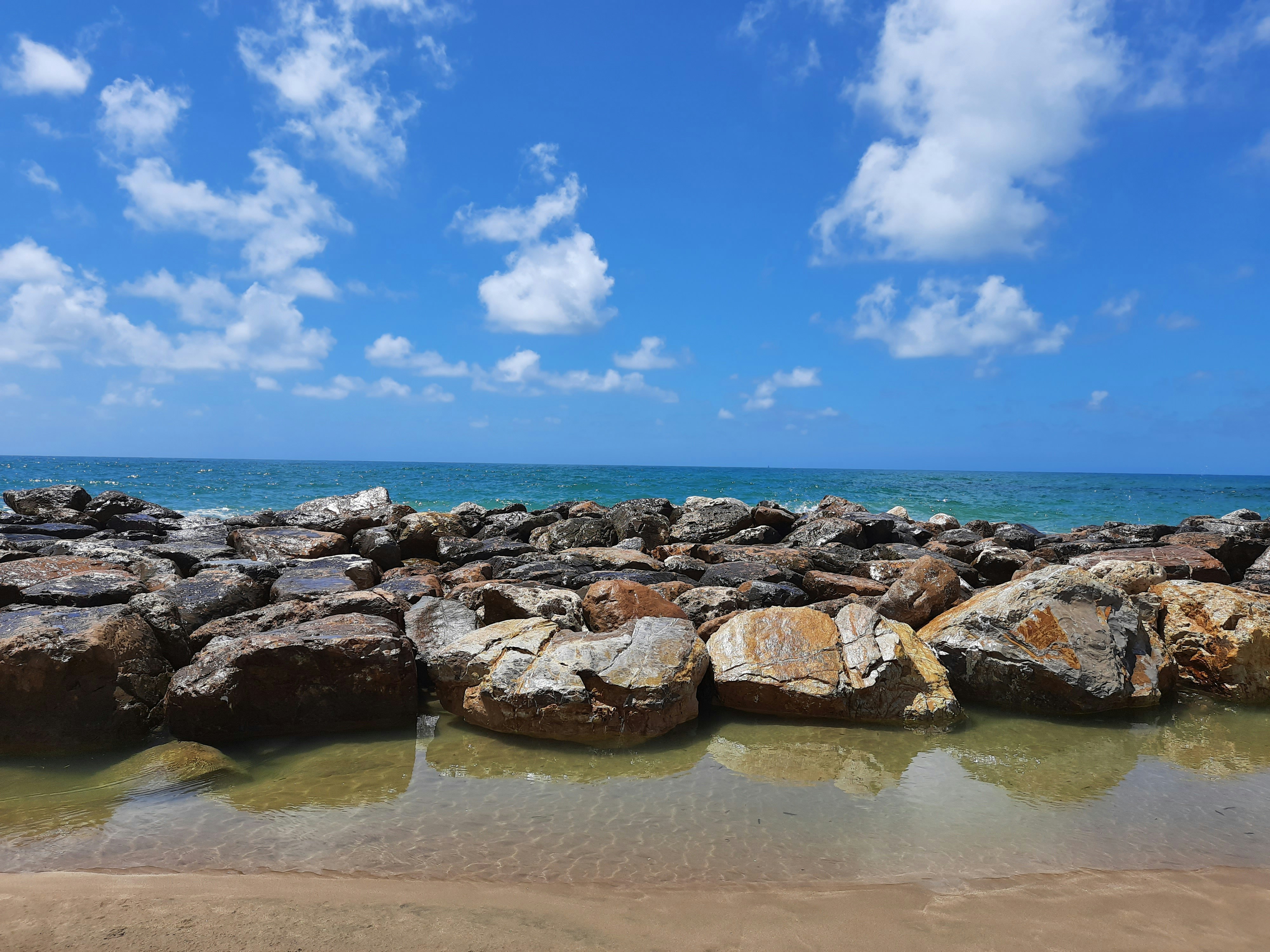 Rocky shoreline with calm waves under a bright blue sky dotted with fluffy white clouds.