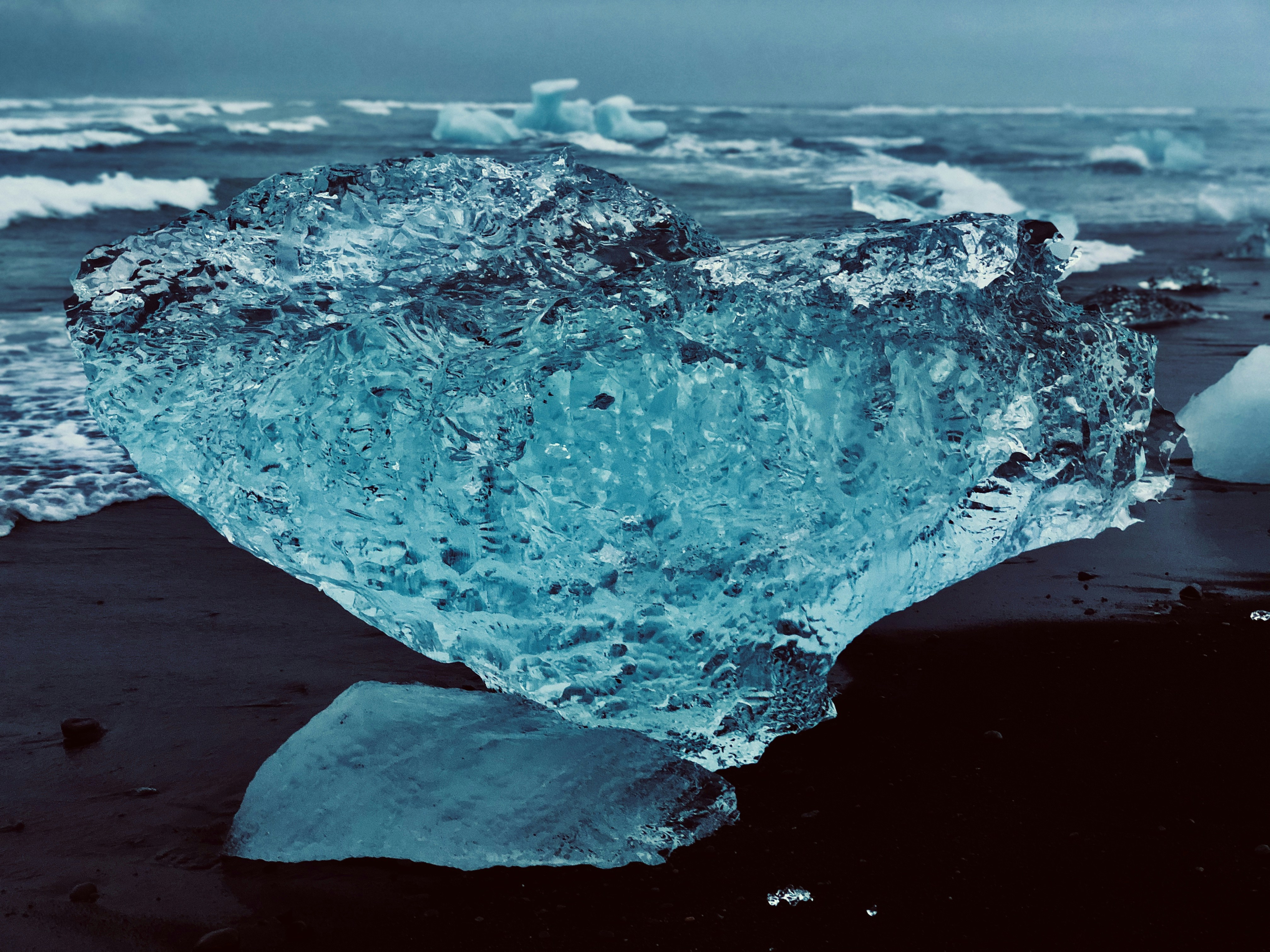 A large, translucent ice formation rests on a black sand beach, contrasting with the turbulent waves in the background.