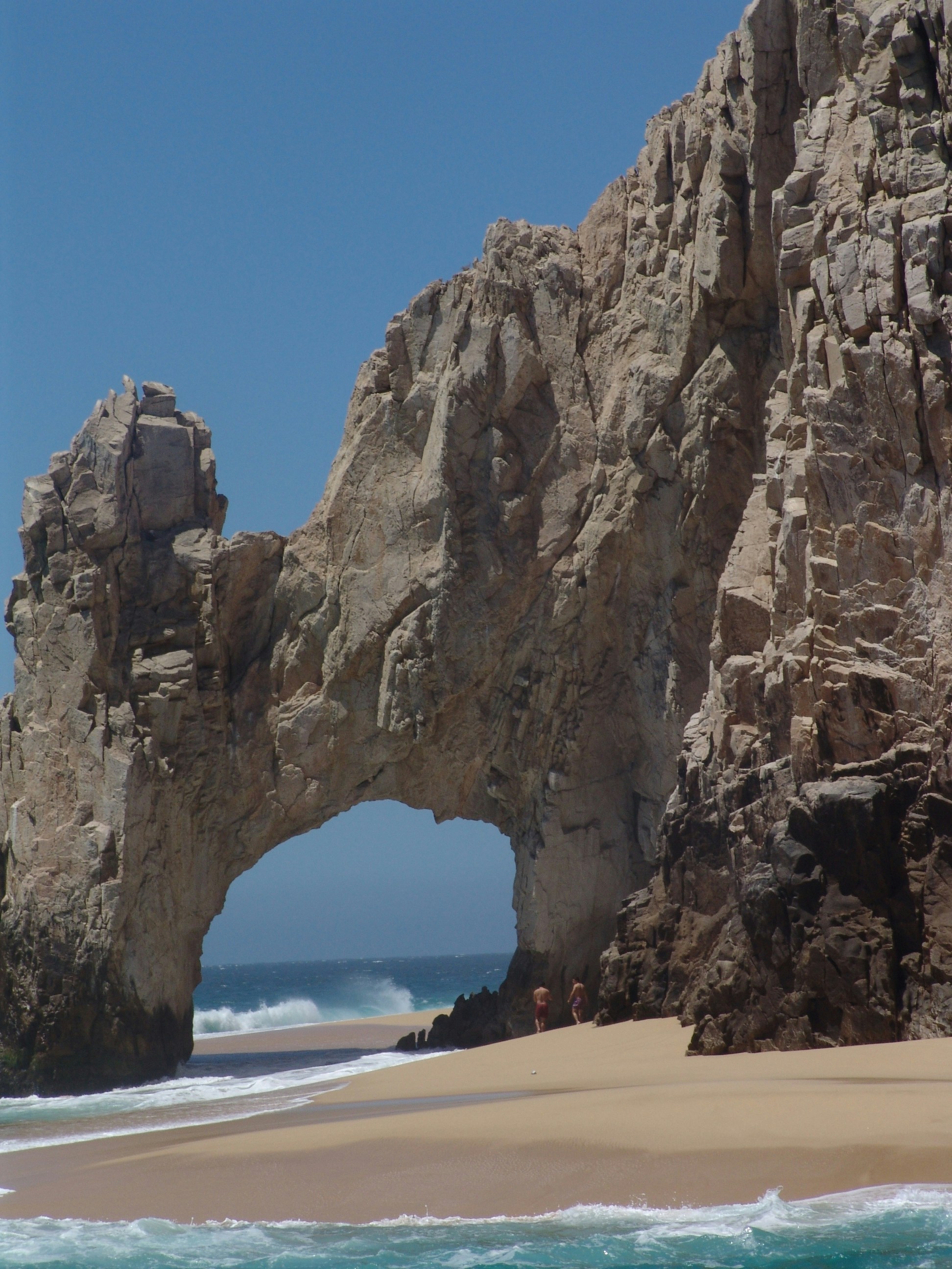 Massive coastal rock arch rises from a sandy beach, framing the turquoise ocean beyond under a clear blue sky.