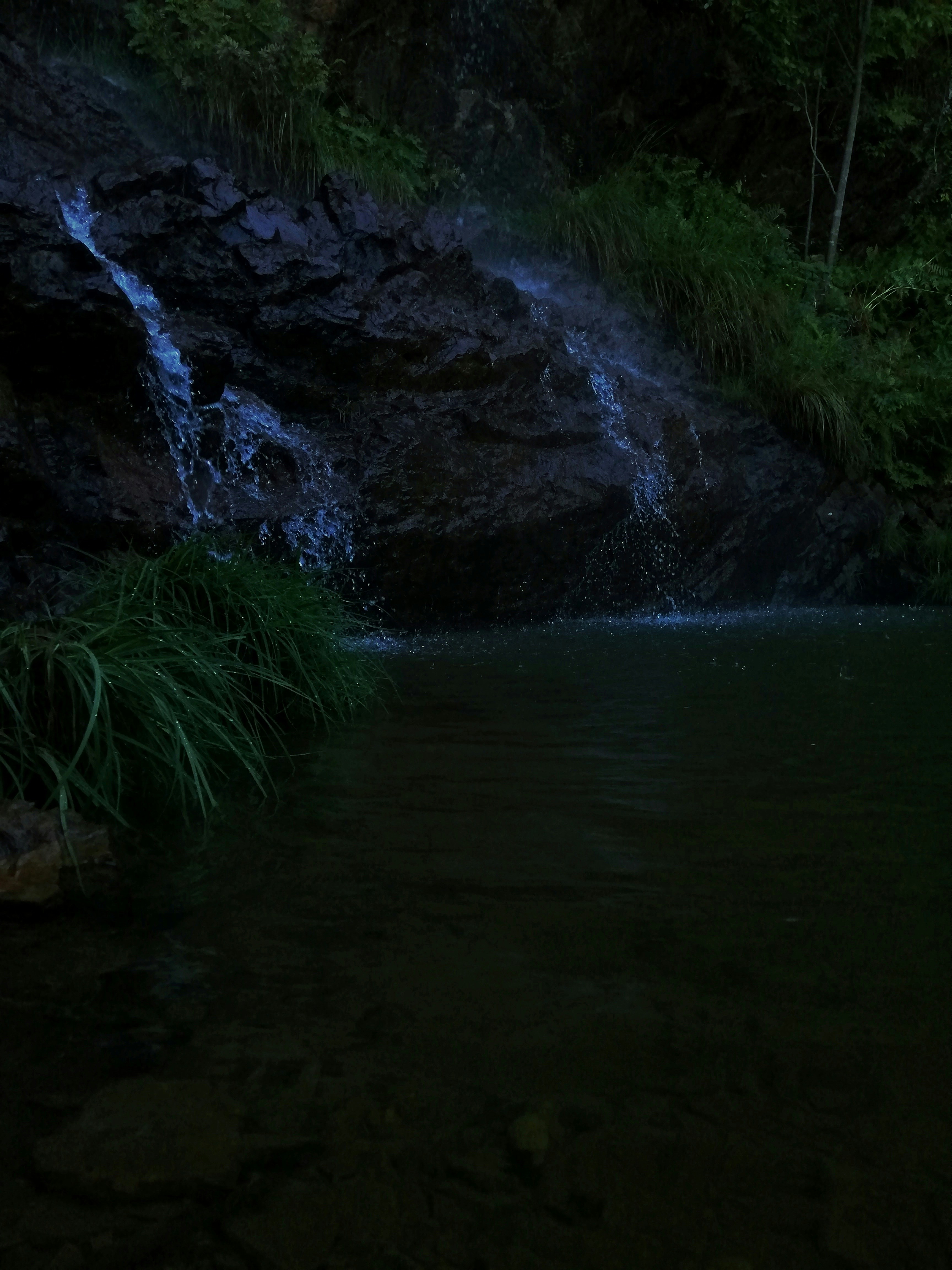 Dim, moonlit cascade spills over dark rock into a quiet pool, with grasses lining the shadowed bank.