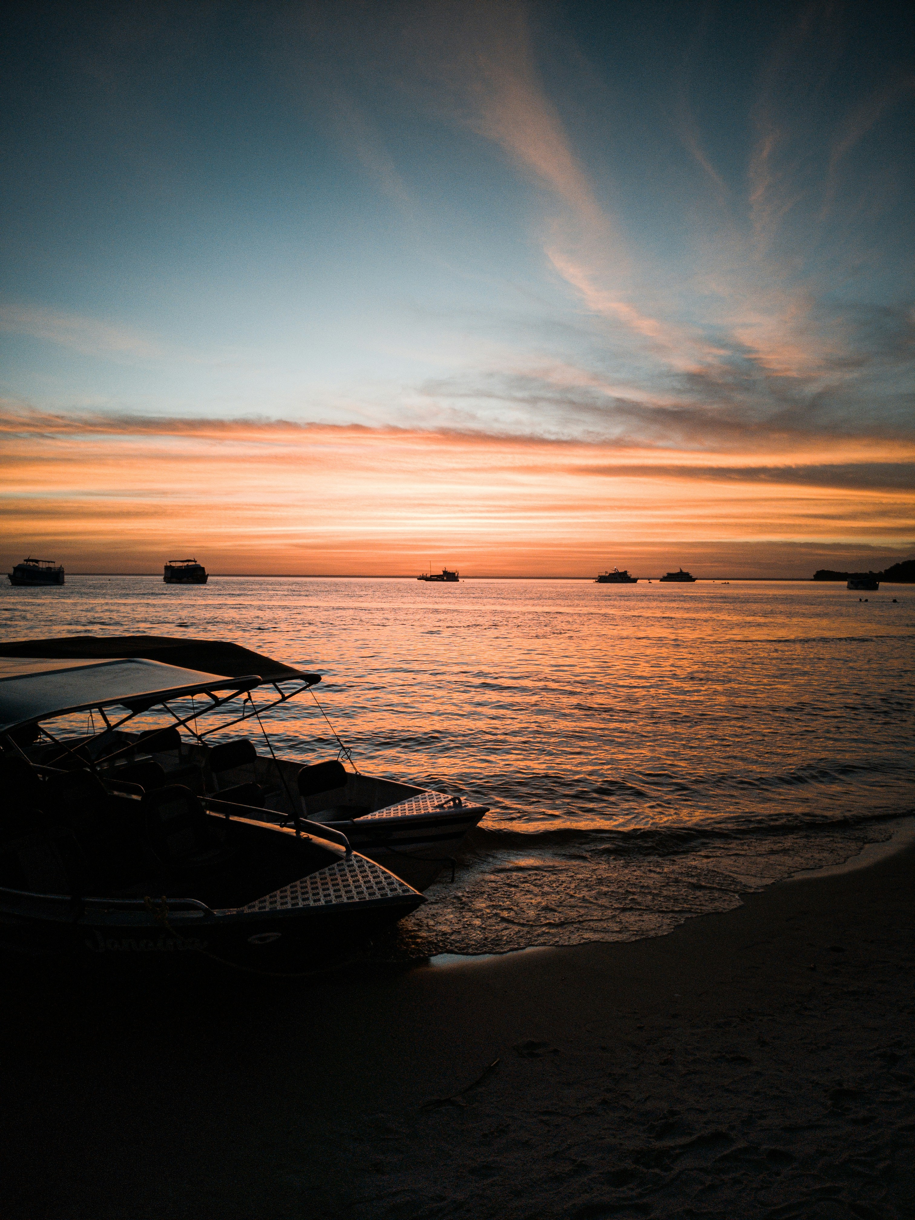 Silhouetted boats resting on a tranquil beach as the sun sets, casting warm hues across the sky and water.
