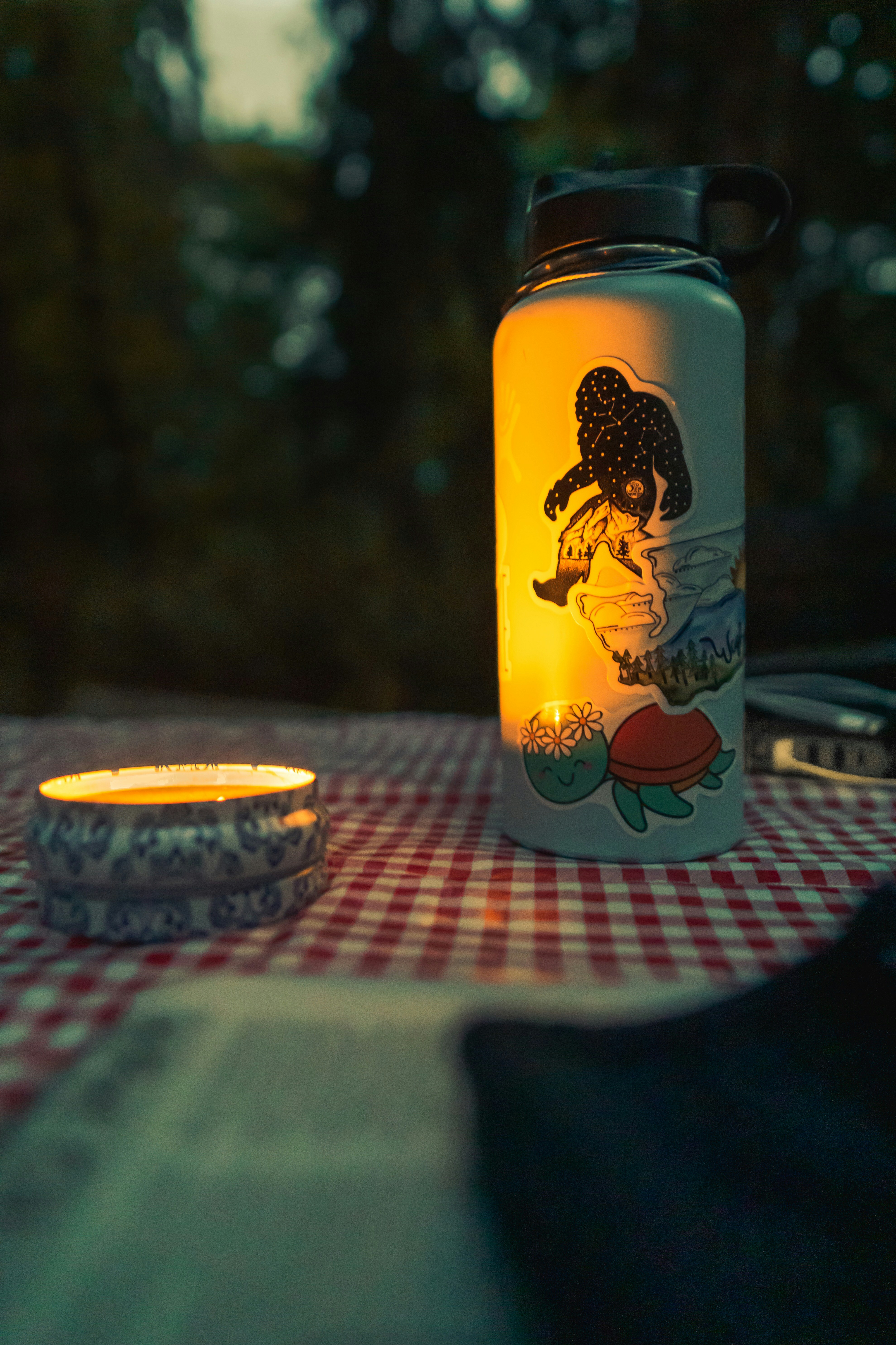 A colorful water bottle illuminated by candlelight, featuring a playful illustration of a turtle and a whimsical figure. The scene captures a cozy outdoor ambiance.