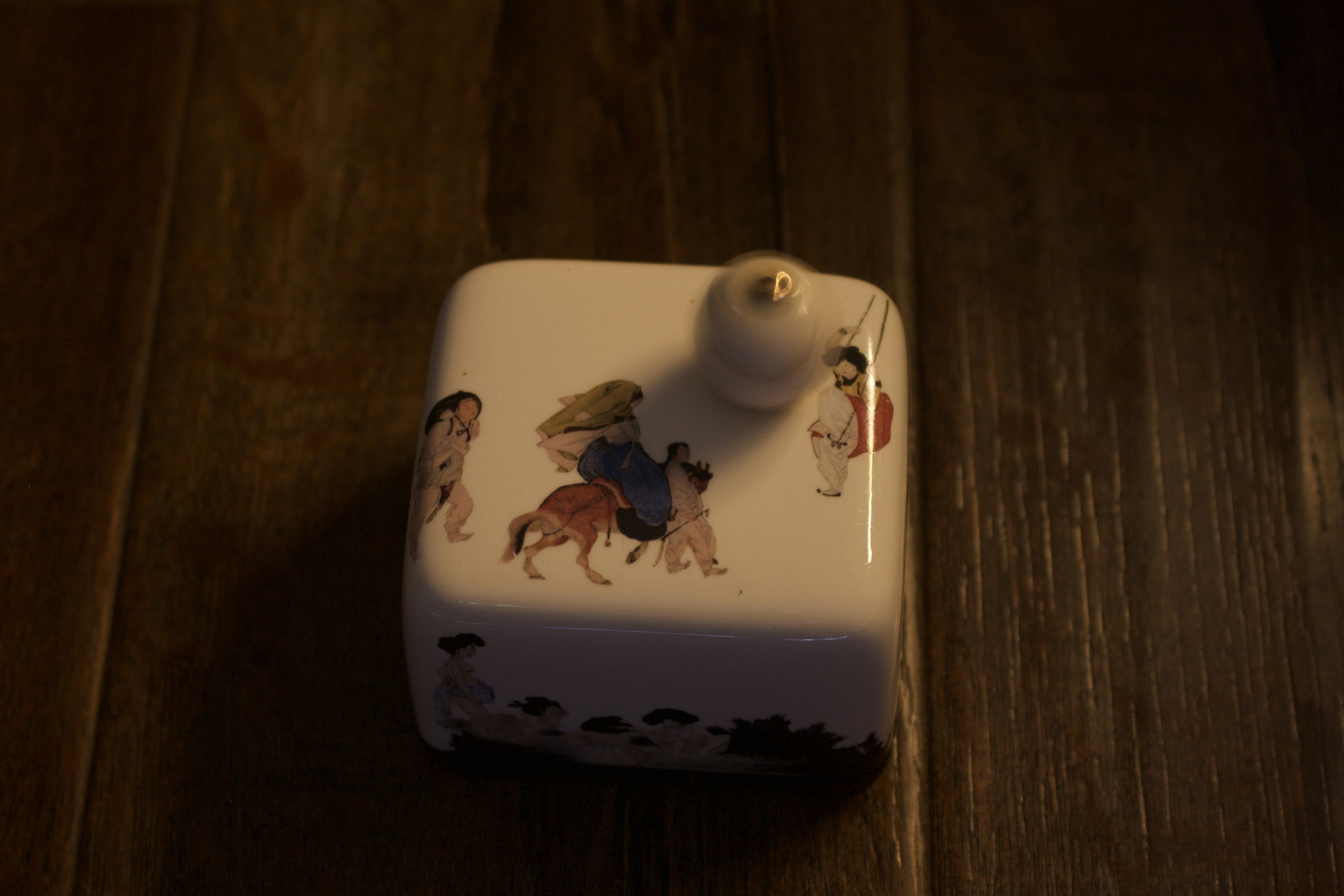 Decorative porcelain box adorned with intricate illustrations of figures and animals, illuminated softly to highlight the craftsmanship.